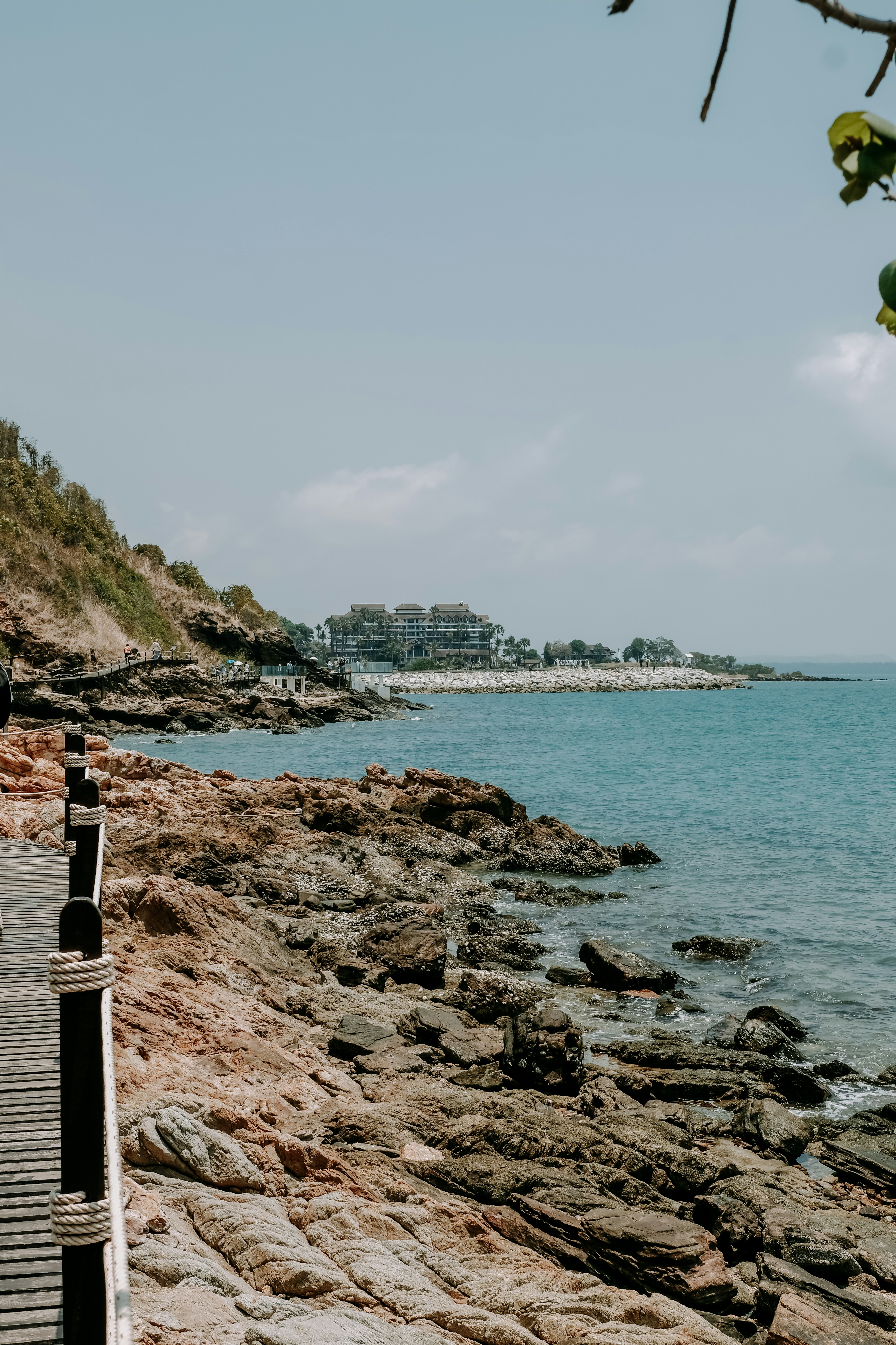 Rugged shoreline path: sunlit rocks, calm turquoise sea, distant cluster of beachfront buildings