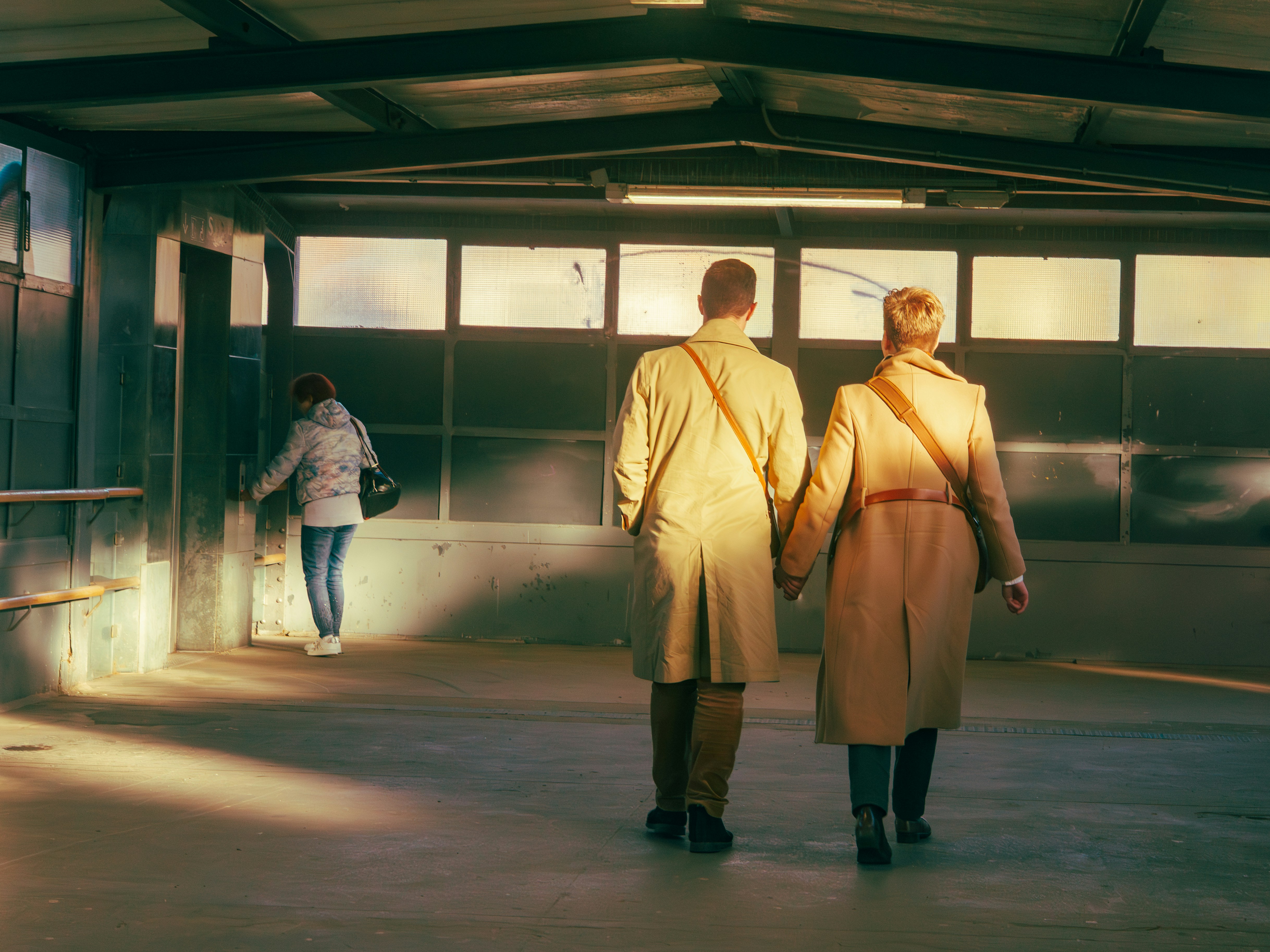 Two figures in trench coats walk hand-in-hand toward a bright exit while a lone person turns toward the wall in a sunlit stairwell