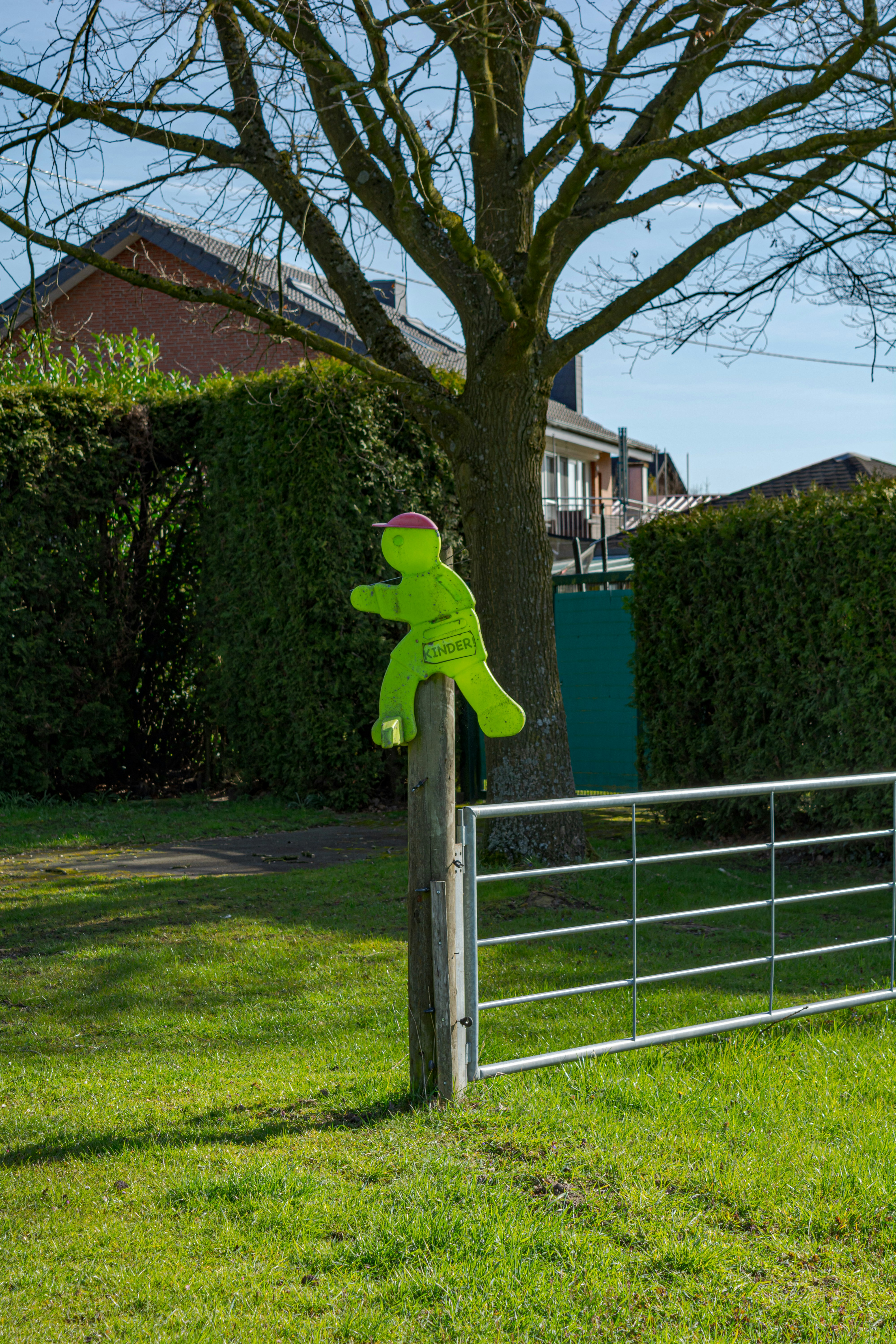 Neon-green playground figure guards the gate against a sunlit hedge