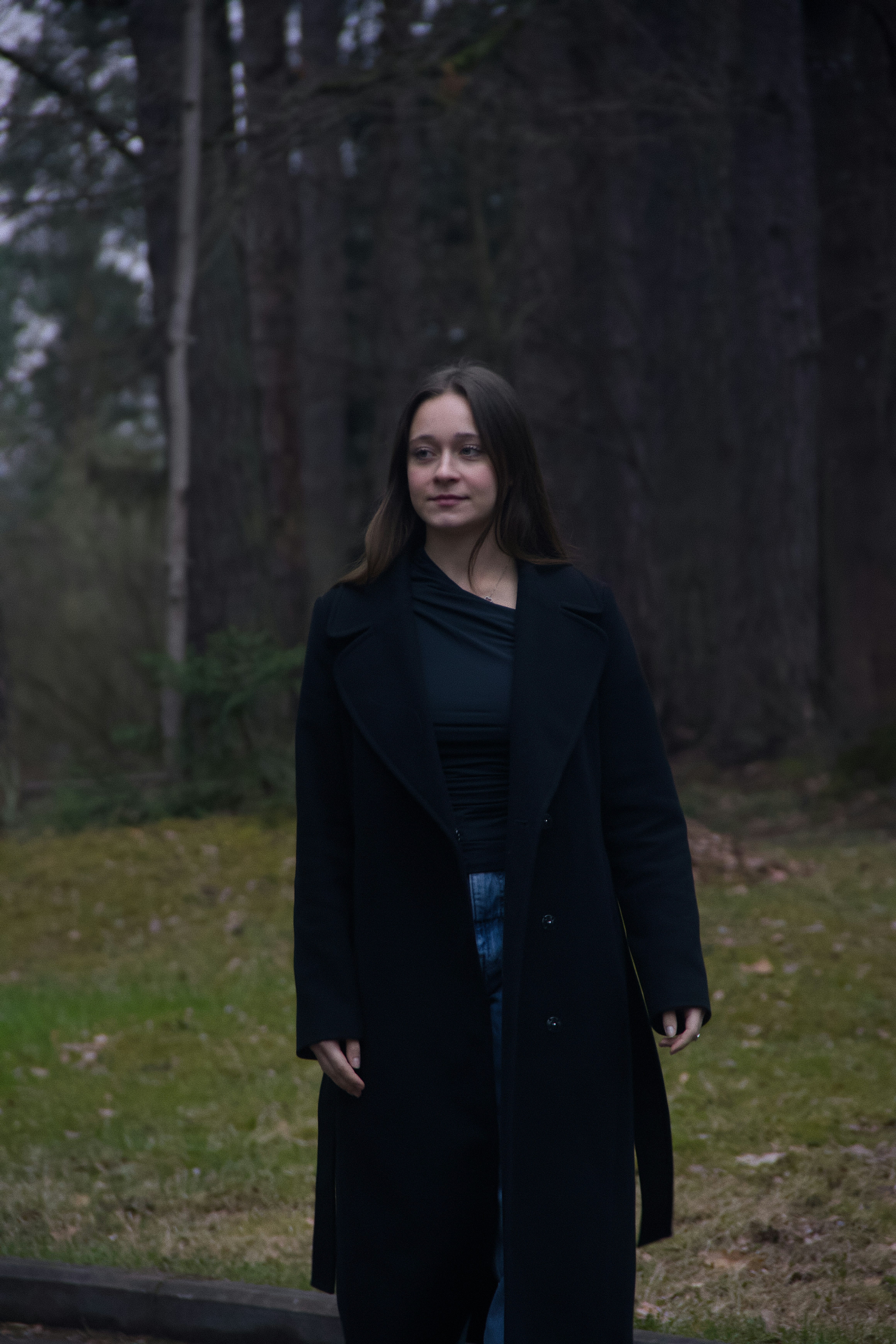 Woman in a dark coat stands softly lit in a forest clearing