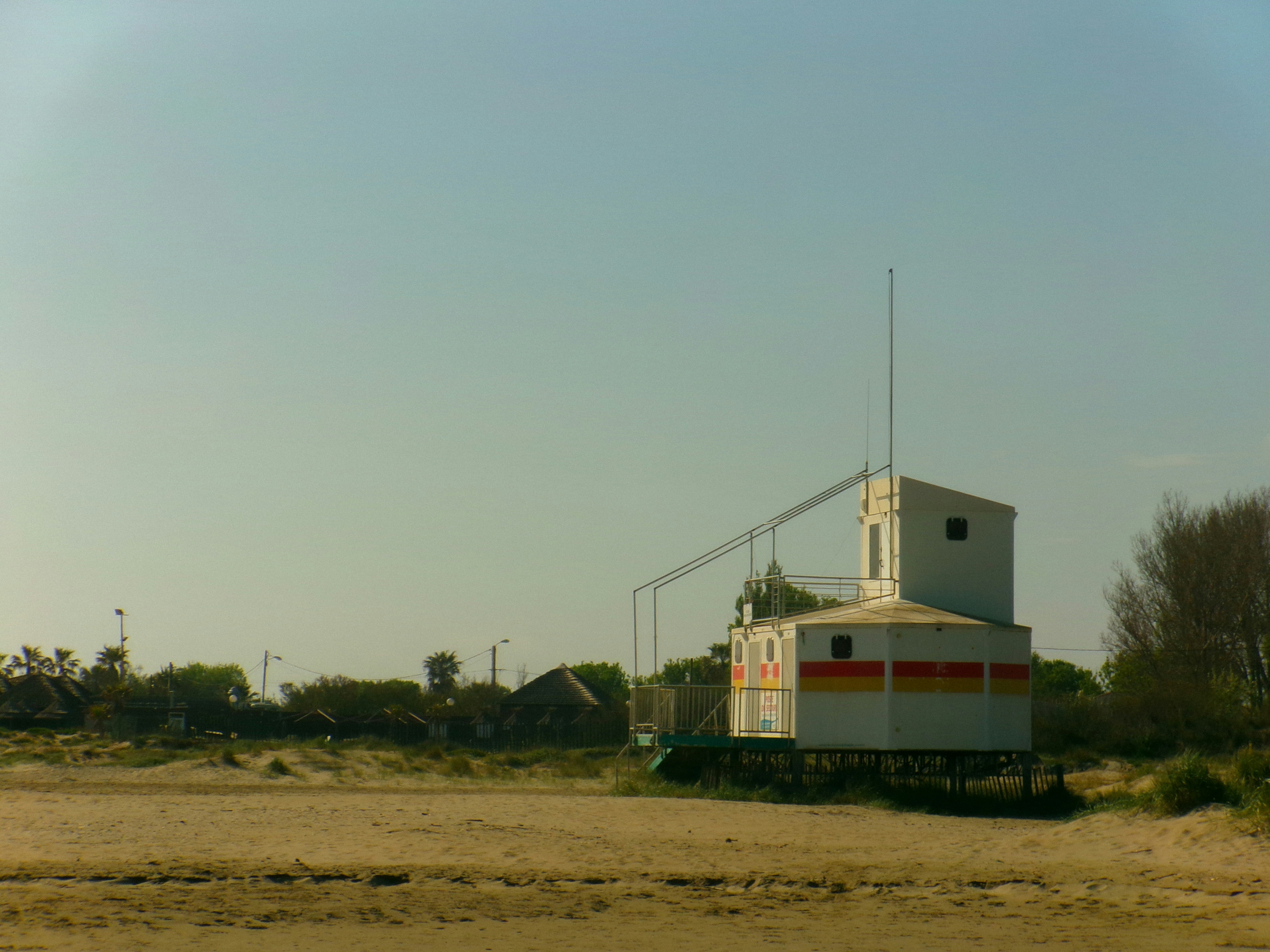 Coastal lifeguard station on a sandy beach with low vegetation and distant dunes, a calm sky overhead.