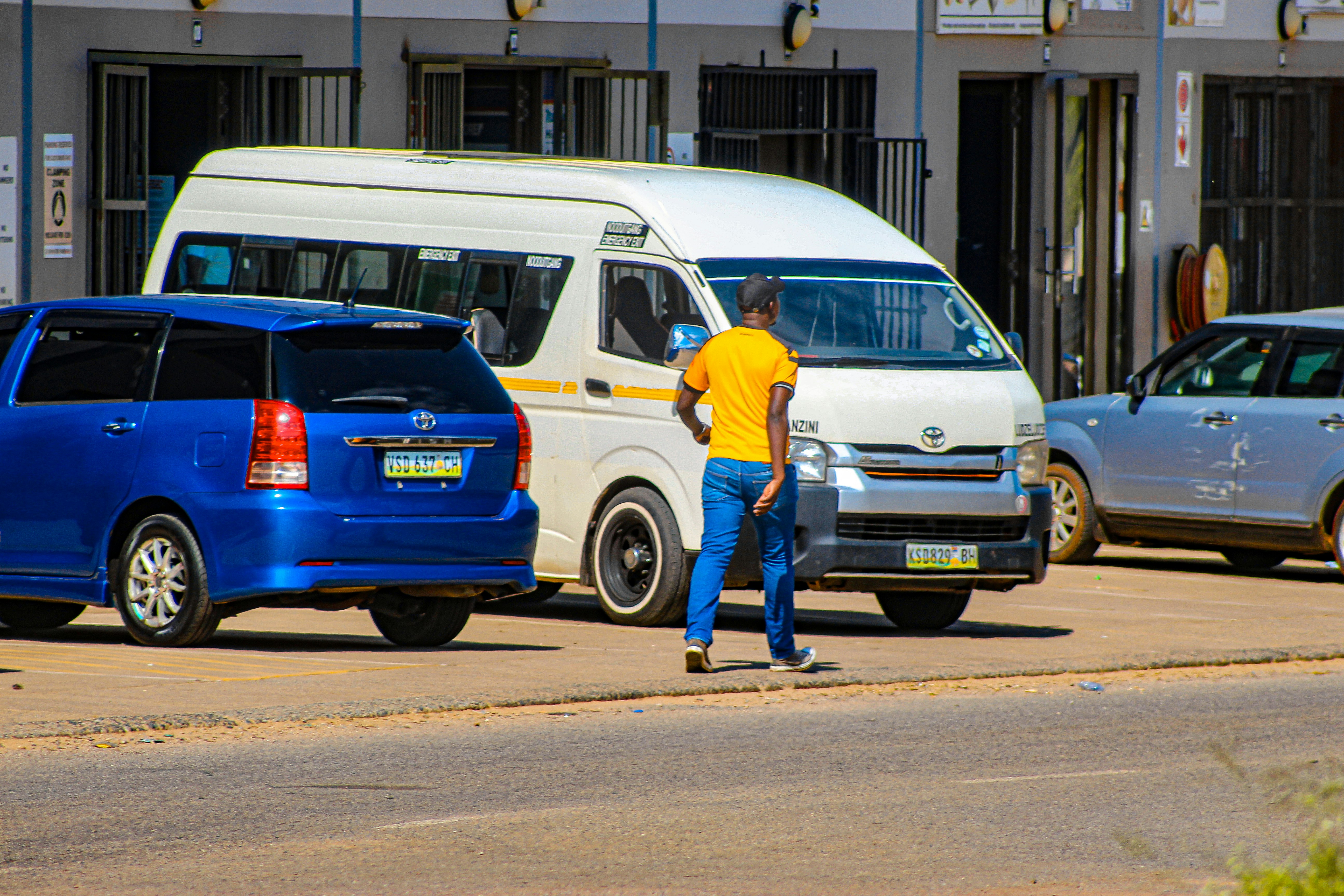 A man stands between a white minibus and blue car.