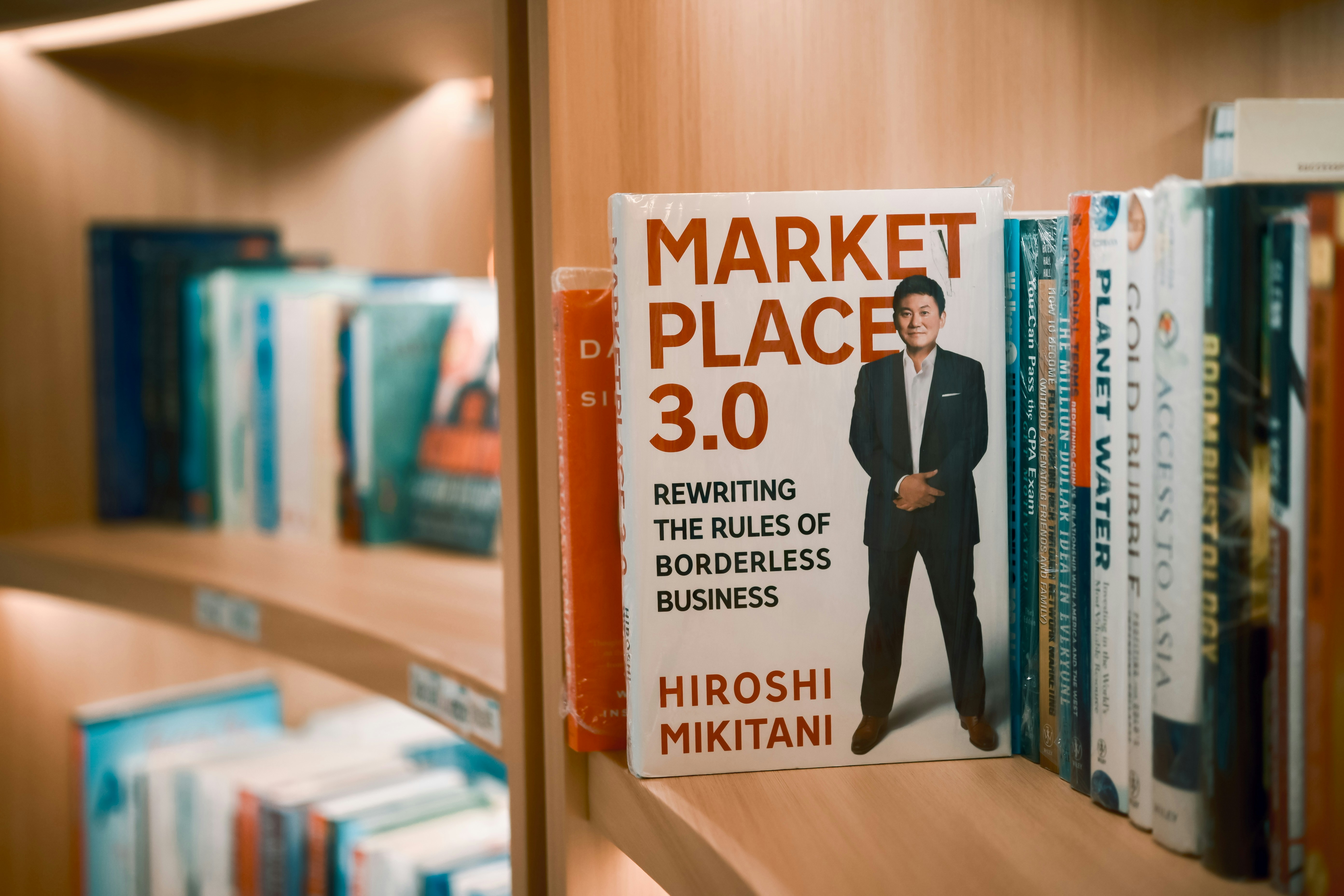 A bookshelf featuring a business book titled 'Market Place 3.0' by Hiroshi Mikitani, with other books lined along the wooden shelf.