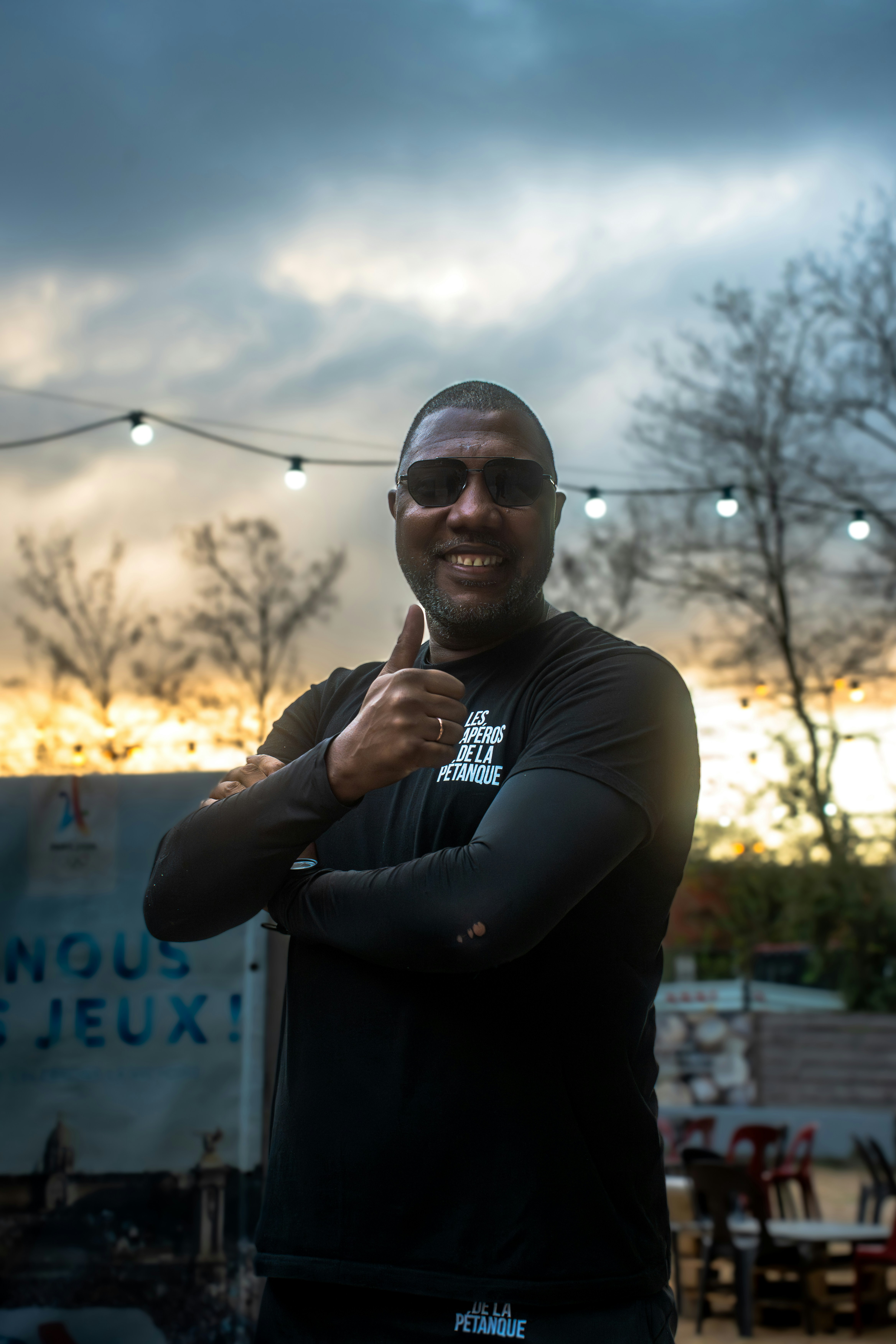 Man in dark athletic shirt gives thumbs up under string lights at an outdoor venue during sunset; the scene captures warm background glow and casual, confident pose.