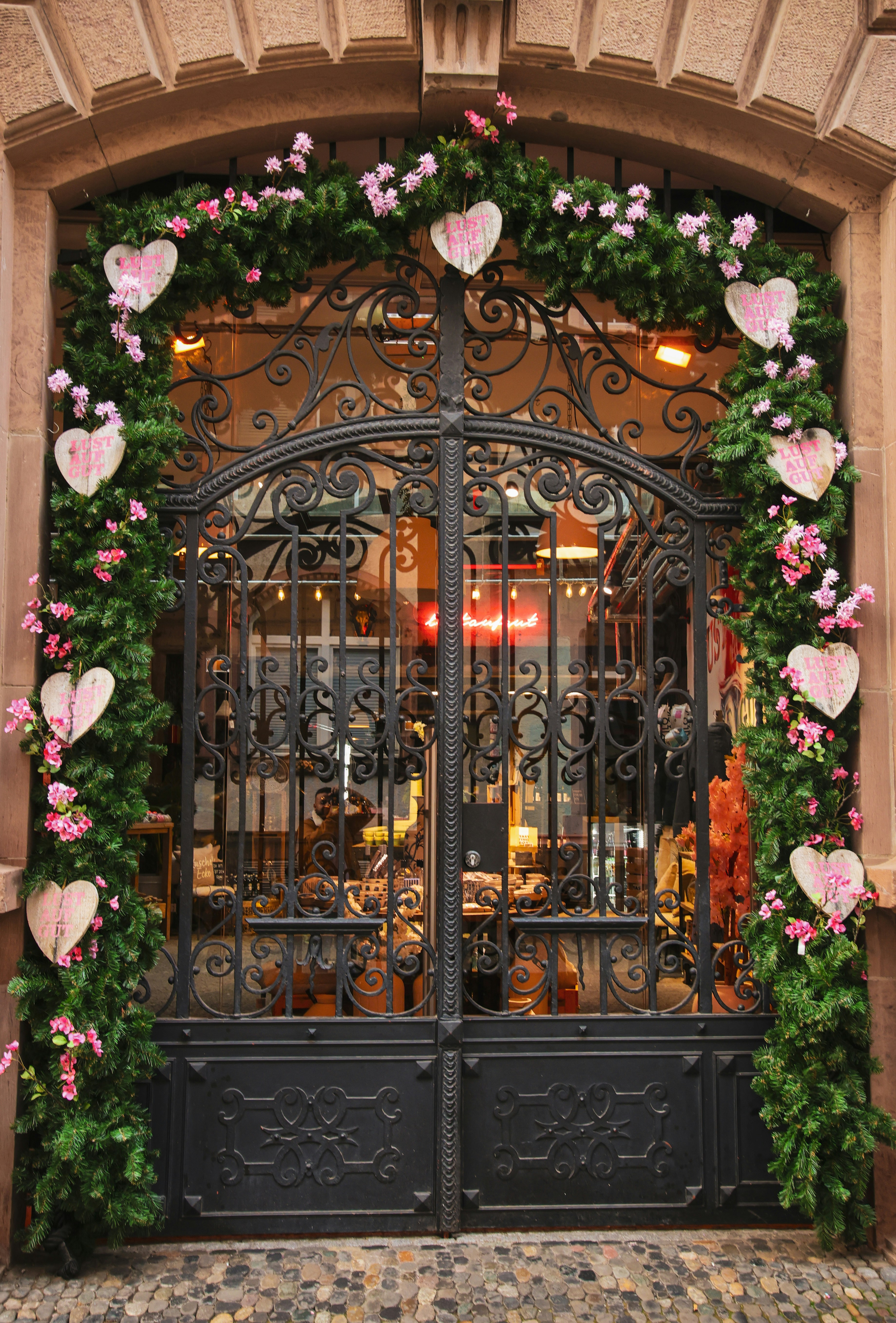 Intricate wrought-iron gate framed by a garland of greenery and pink flowers with heart shapes, revealing a warmly lit interior through the bars. A decorative storefront or doorway with ornate craftsmanship.