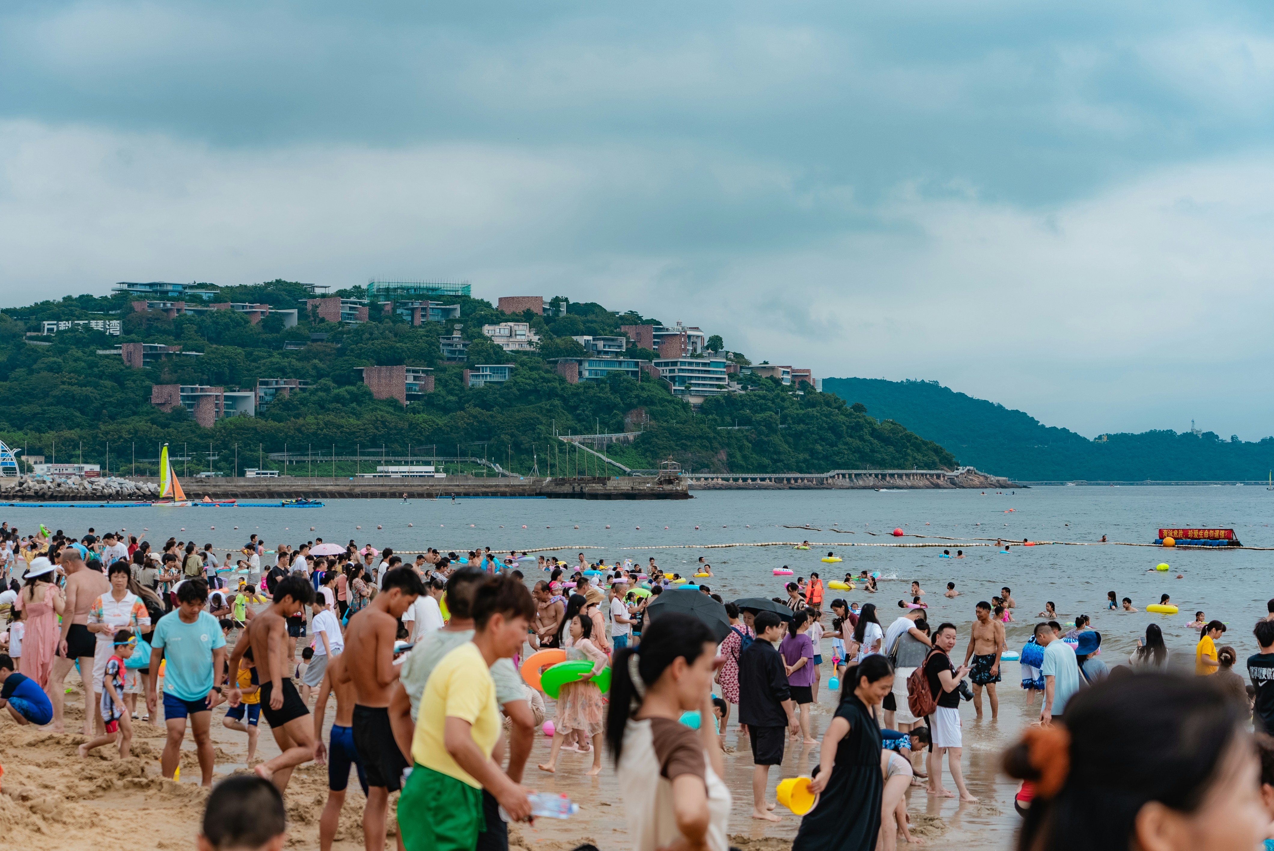Bustling beach scene with numerous people enjoying a sunny day along the shore, with a hillside community in the background.