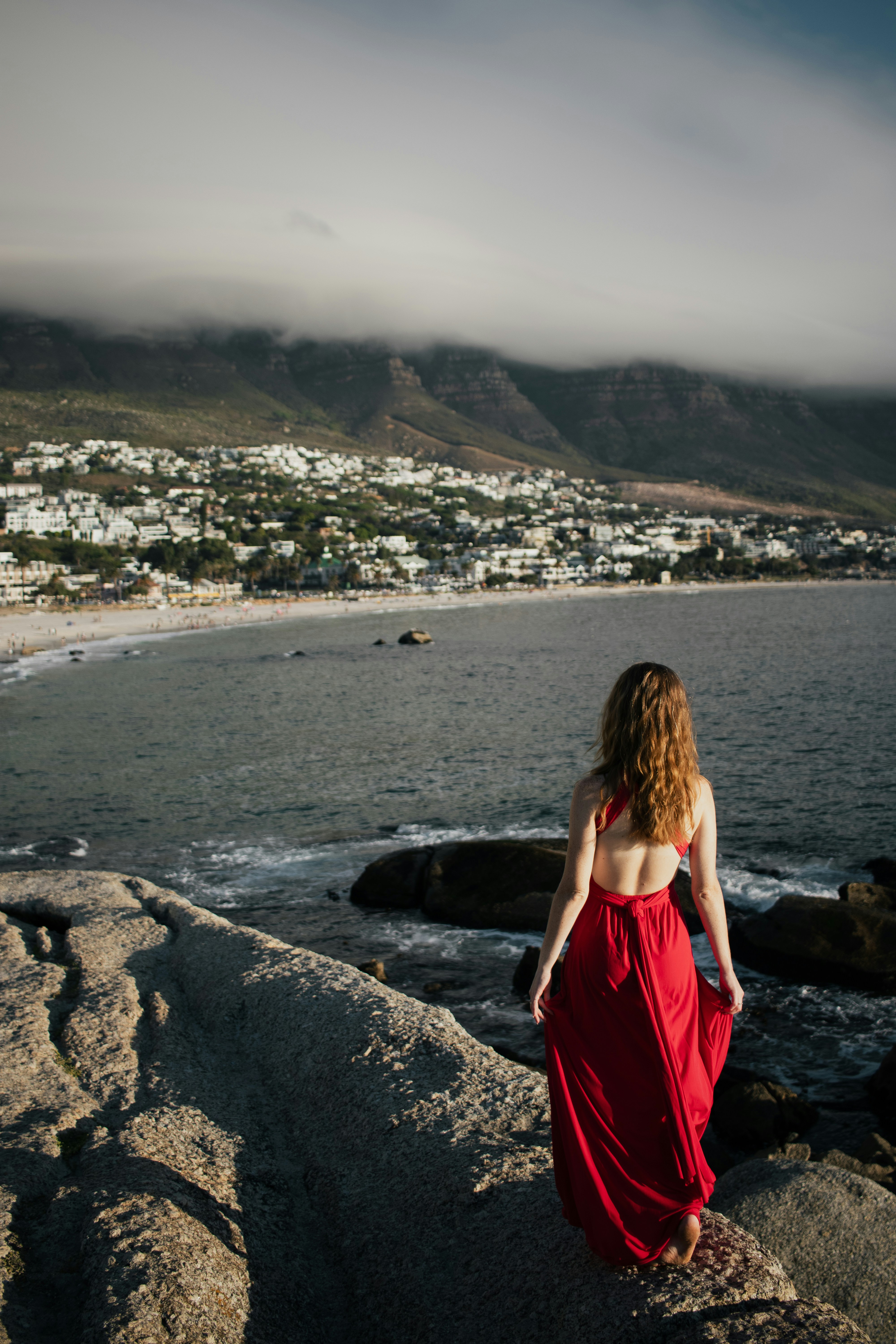 Red Dress at the Edge: A Woman Standing on Coastal Rocks with a Town and Mountains Shrouded in Cloud