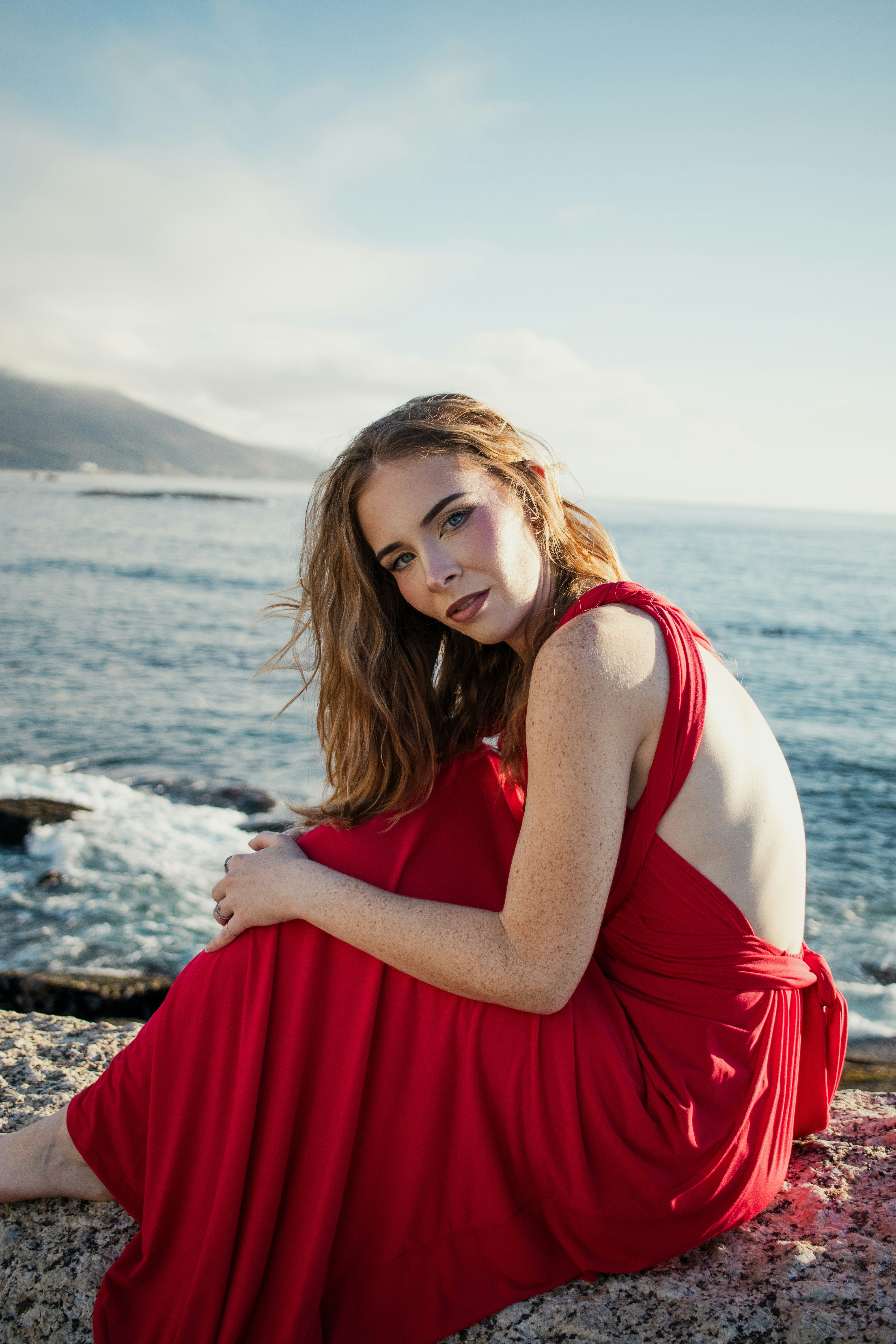 Woman in red dress sits by the ocean.