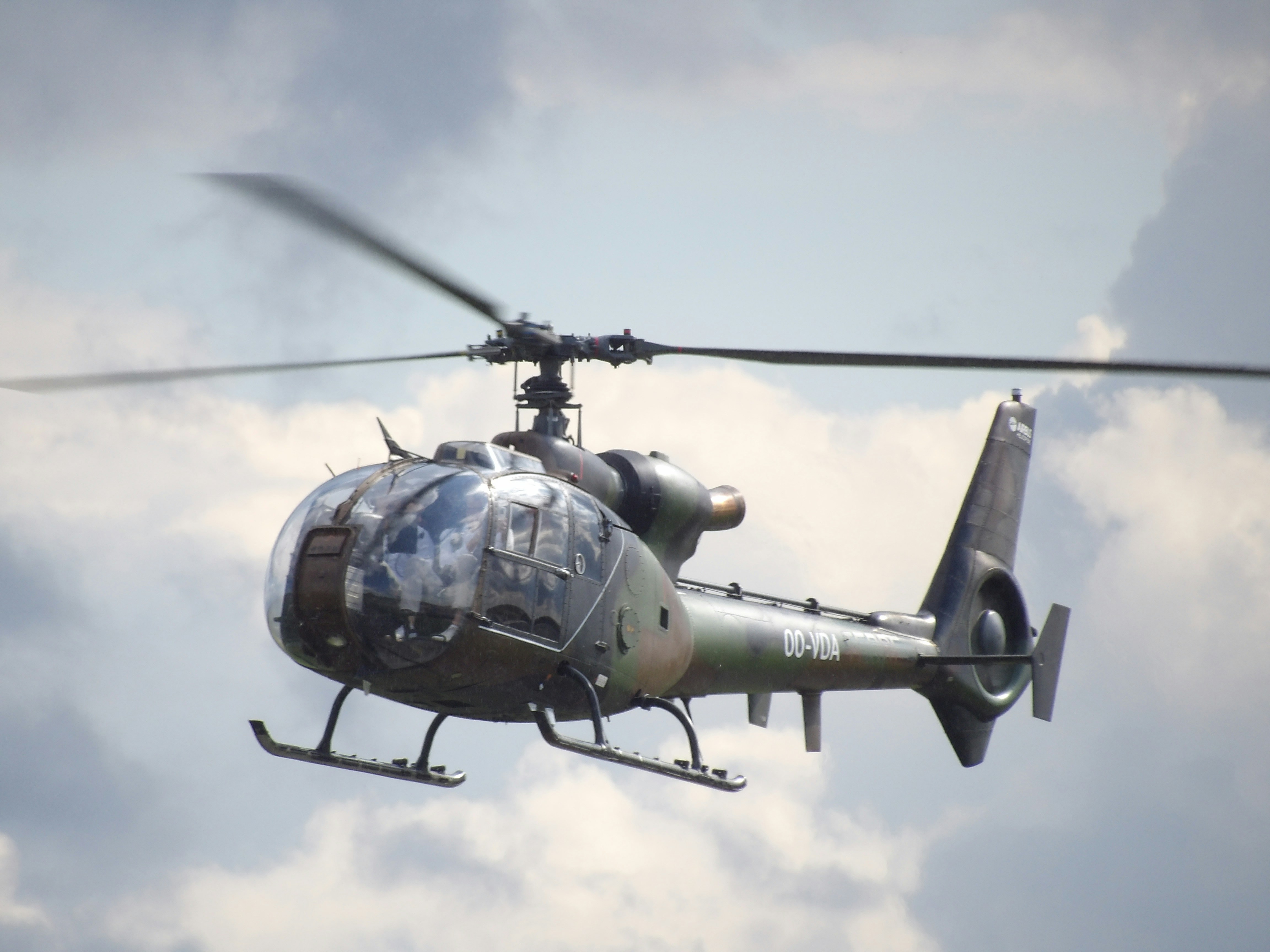 Camouflaged helicopter in flight against a cloudy backdrop, rotor blur indicating motion.