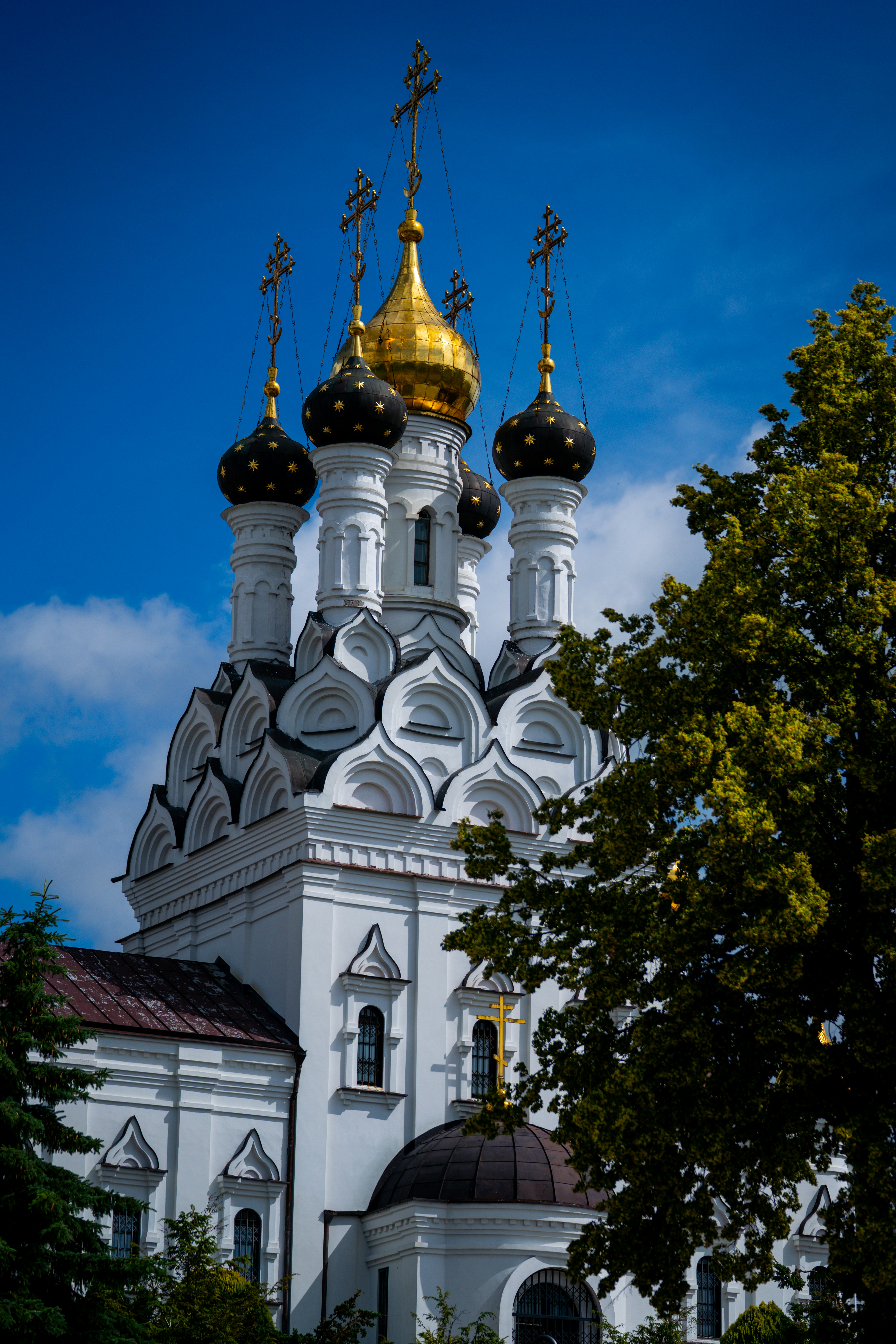 Golden onion domes rise above a white church framed by green leaves under a blue sky