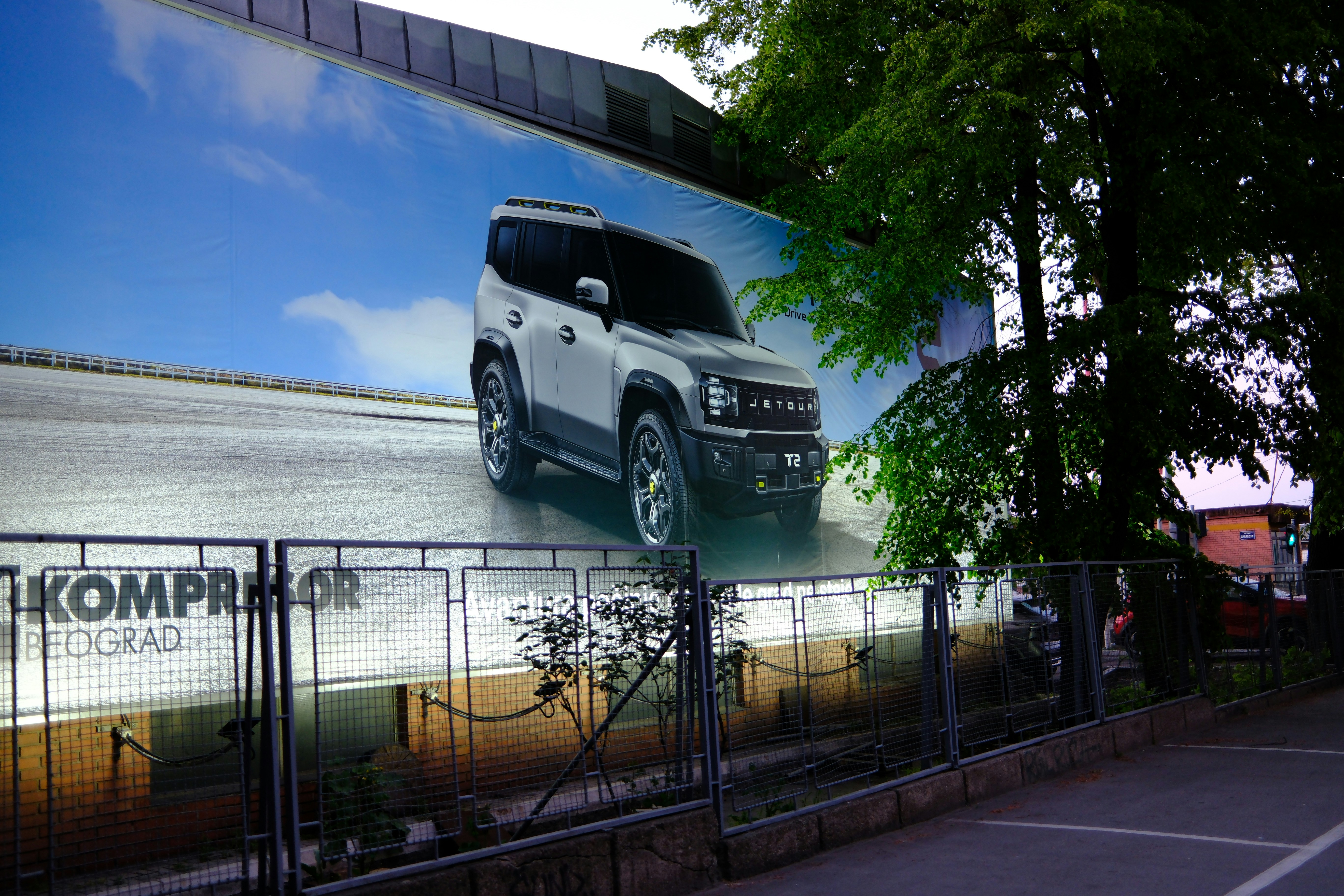 A compact SUV is displayed on a large outdoor banner along a fenced urban wall, with trees and pavement in the foreground.