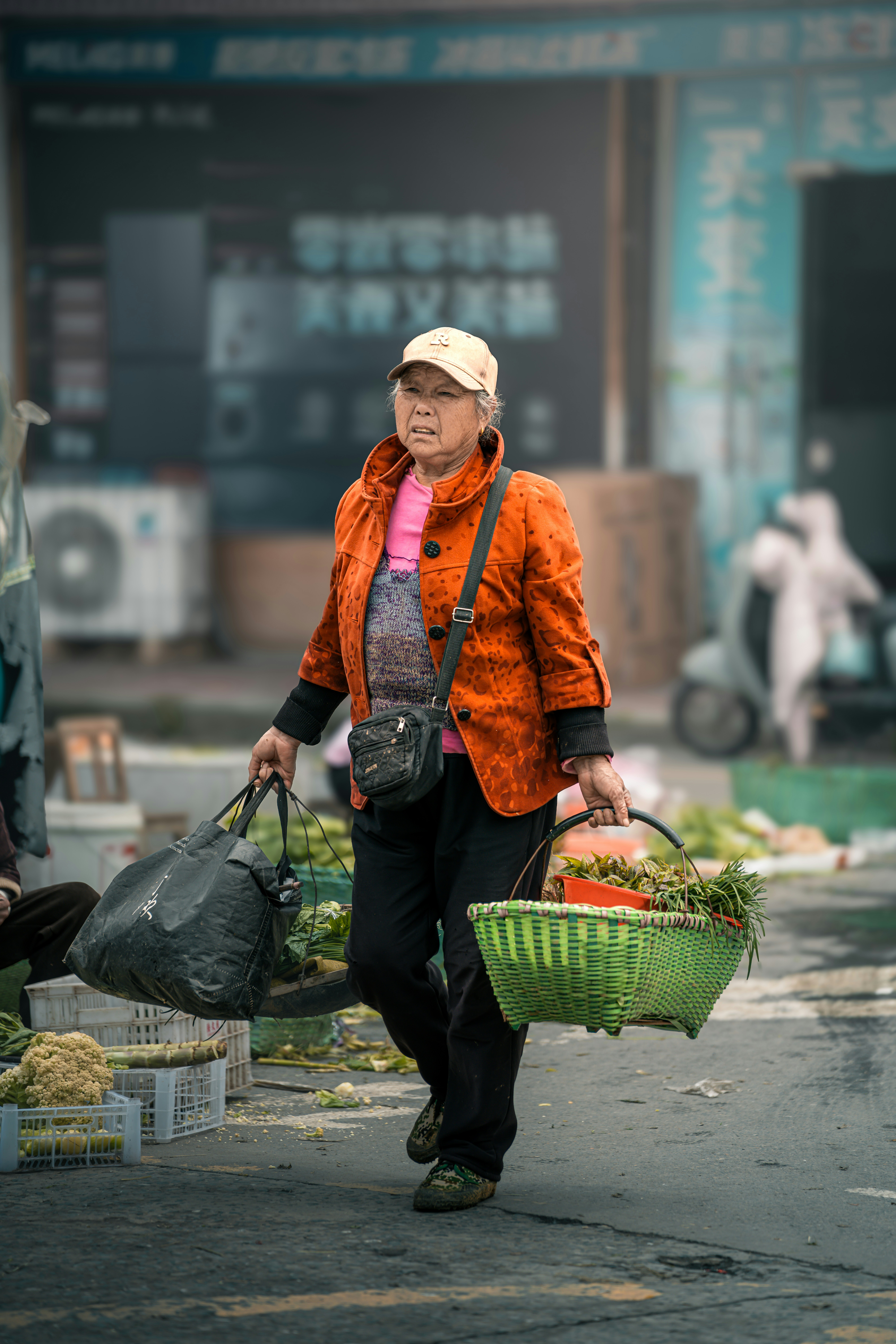 Market scene featuring an elderly woman in an orange jacket carrying a green woven basket and black bag amid urban stalls.