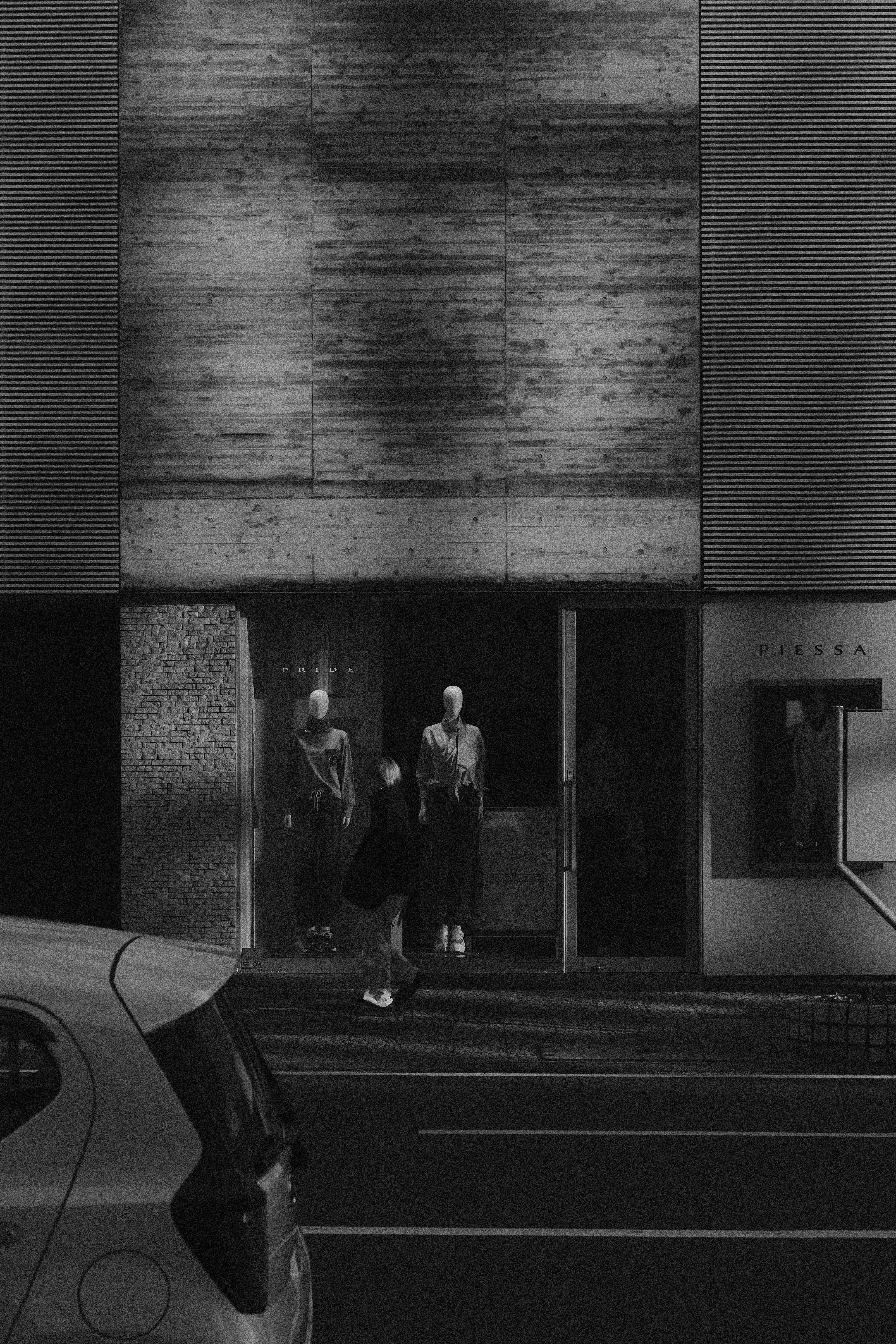 A grayscale street scene shows a storefront with mannequins inside; a pedestrian with a bag walks past, while a car edges the frame. The image emphasizes urban minimalism and reflective textures.