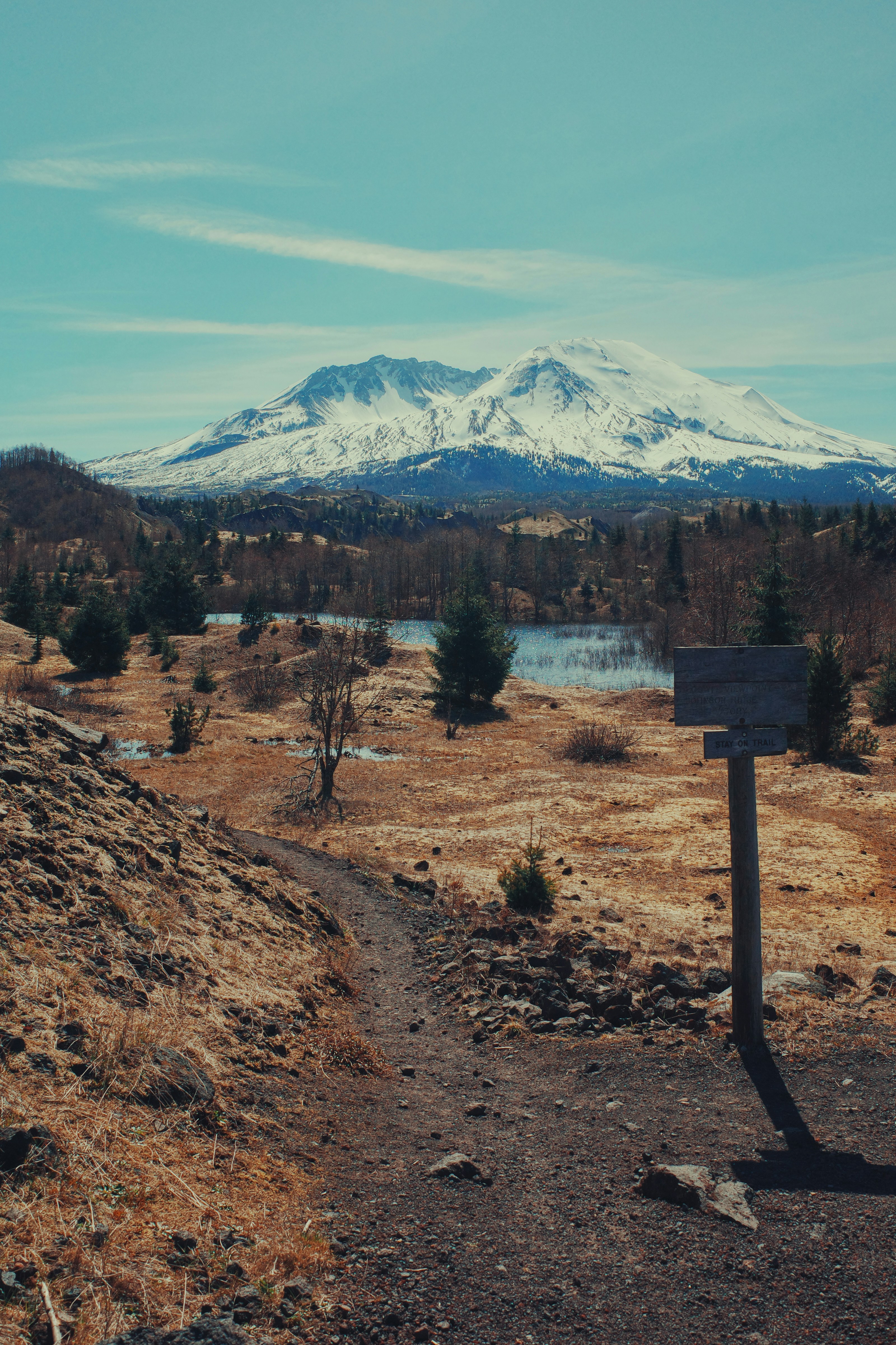 Snow-capped mountains rise above a dry, rocky trail dotted with sparse trees and a lone wooden sign; a tranquil landscape capturing distant alpine scenery.