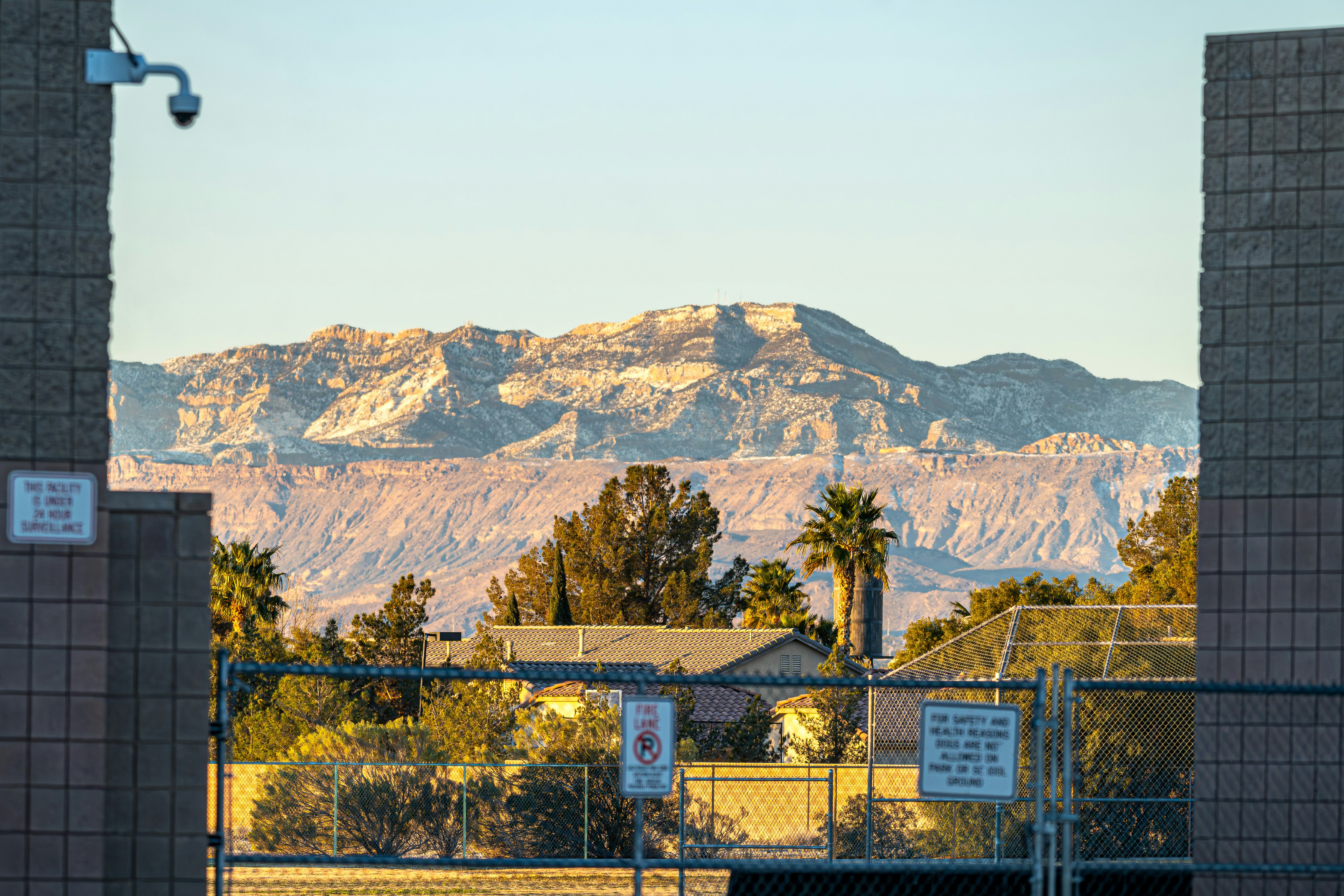 Distant desert mountains rise behind a chain-link fence and urban foreground, framed by two brick columns under clear daylight.