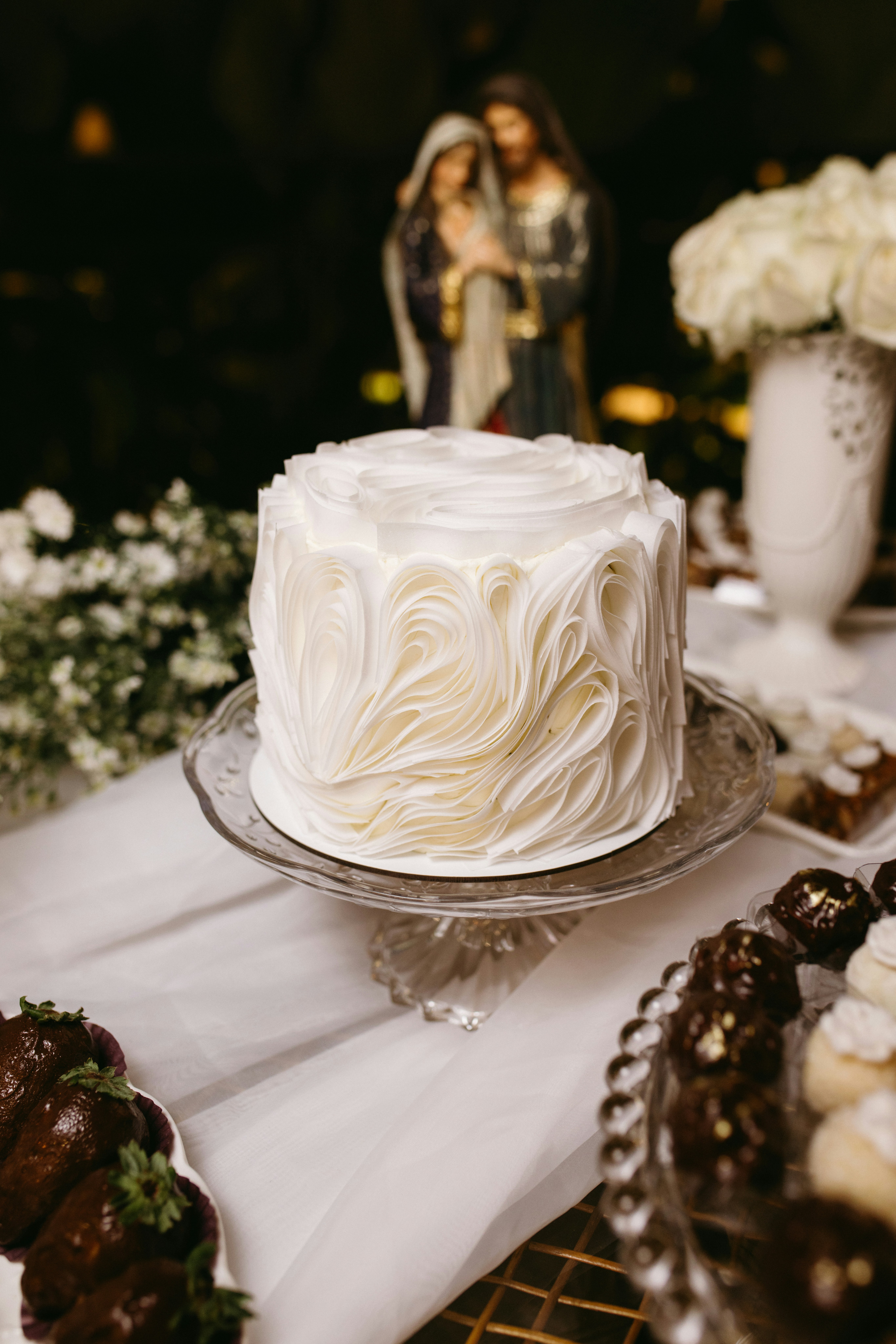 White Rosette Cake on a Glass Stand with Blurred Wedding Couple in the Background
