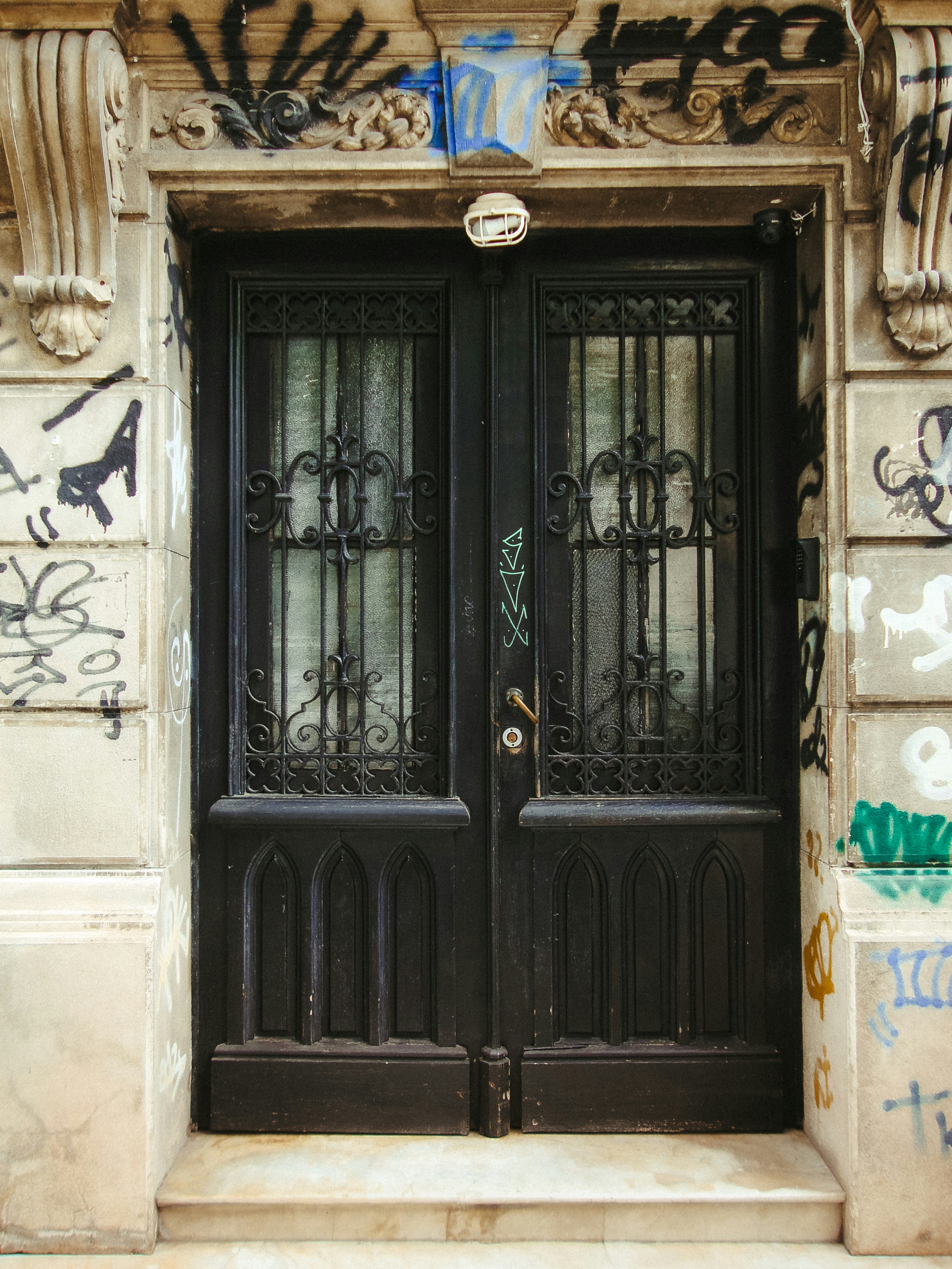 Dark mirrored doors with intricate ironwork sit within a graffiti-tagged stone facade, inviting entry and hinting at urban history.