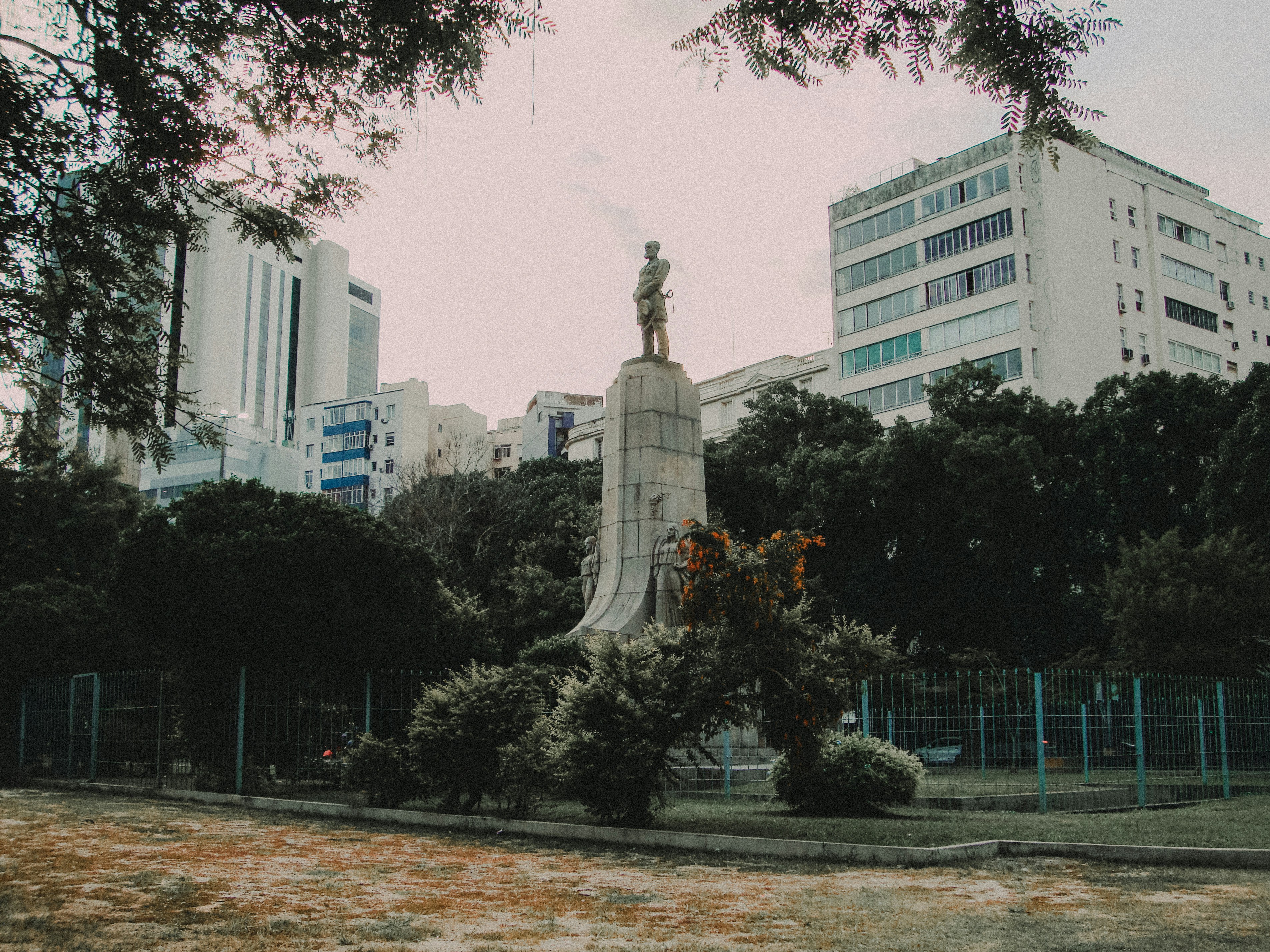 Statue atop a pedestal surrounded by trees and buildings in a city park.