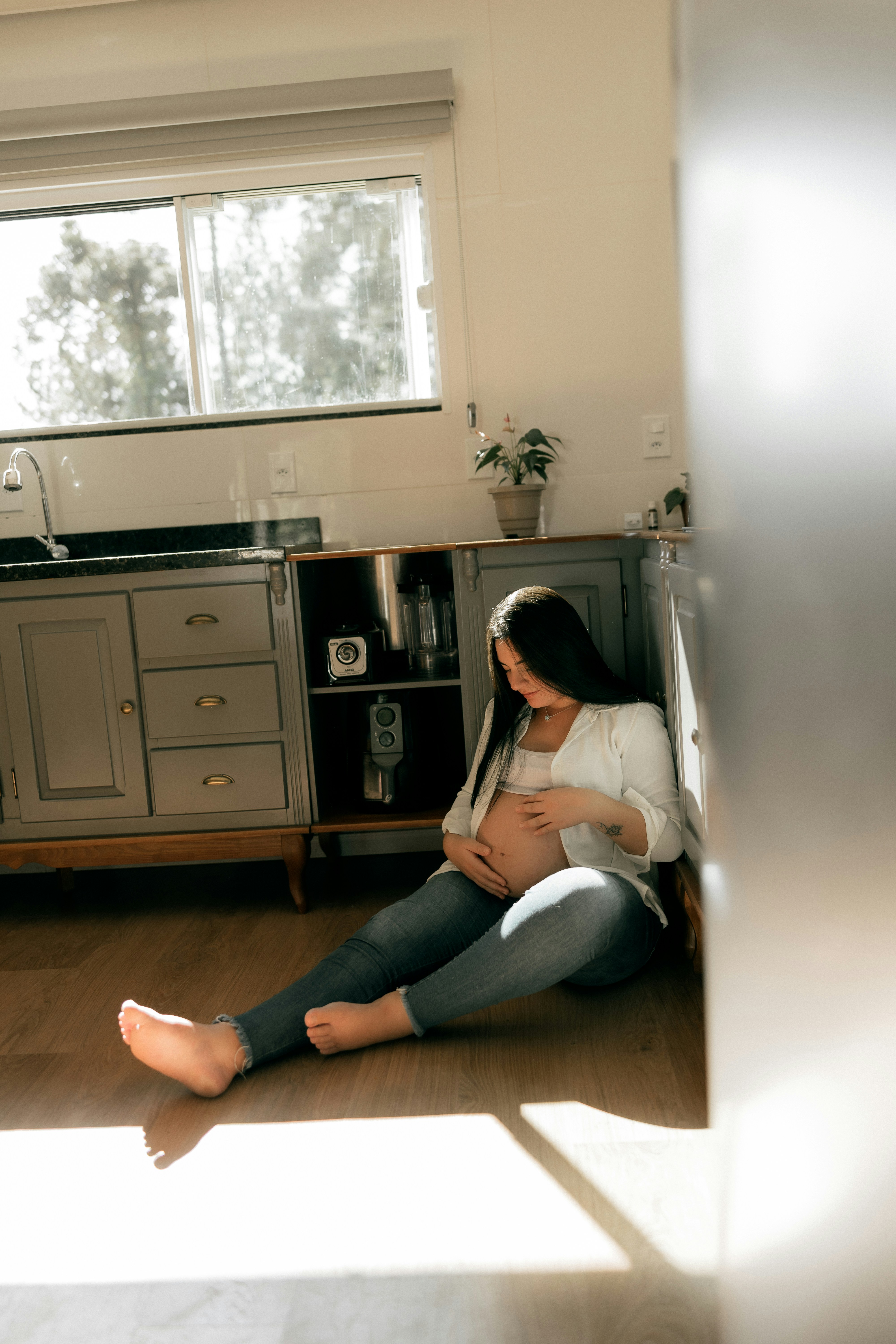 Soft morning light spills across a pregnant woman seated on the kitchen floor, hands resting on her belly. The scene captures a calm, intimate maternity moment in a modern home.