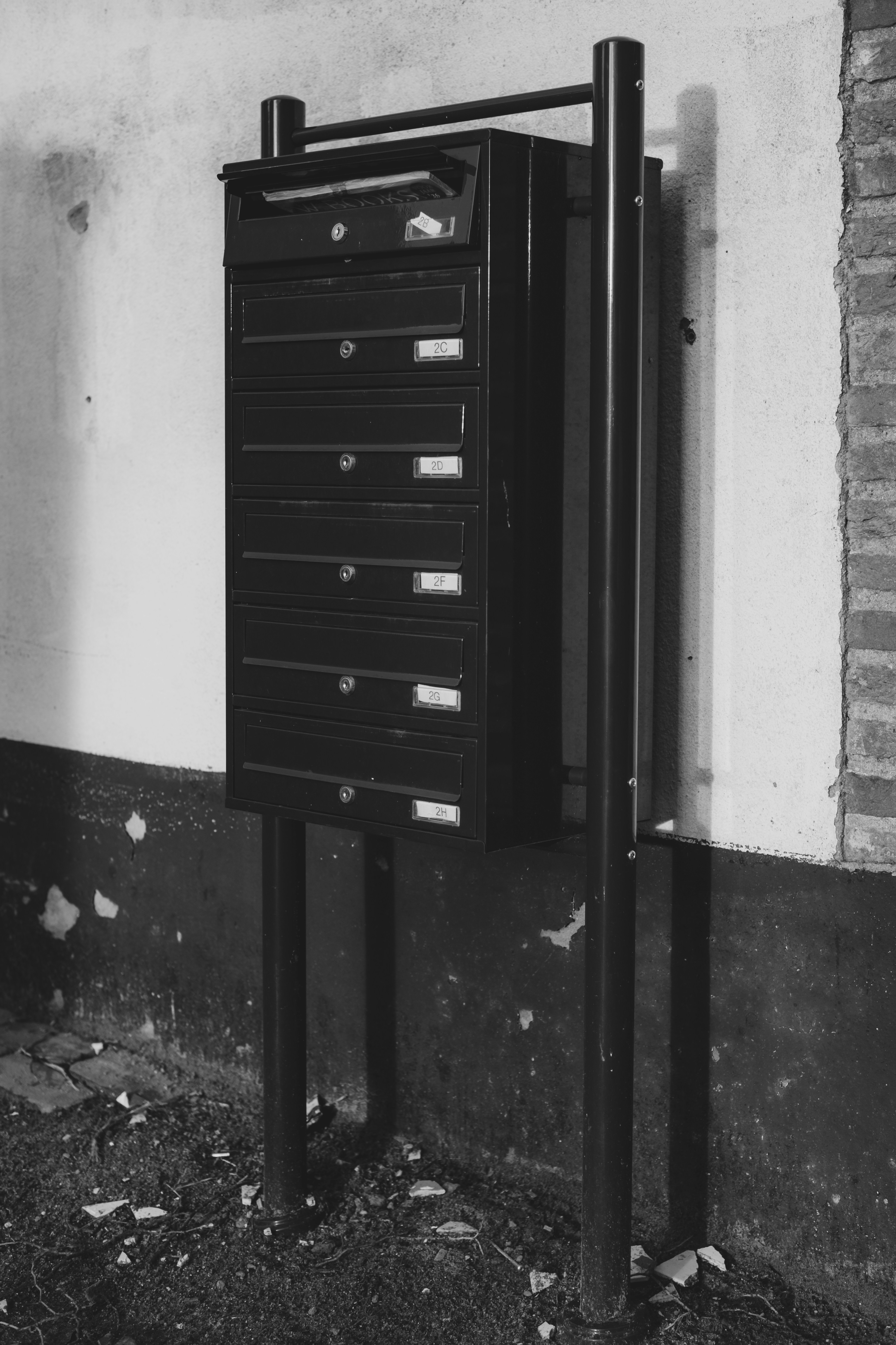 A row of vertical mailboxes mounted on black posts sits against a weathered wall, captured in black and white.