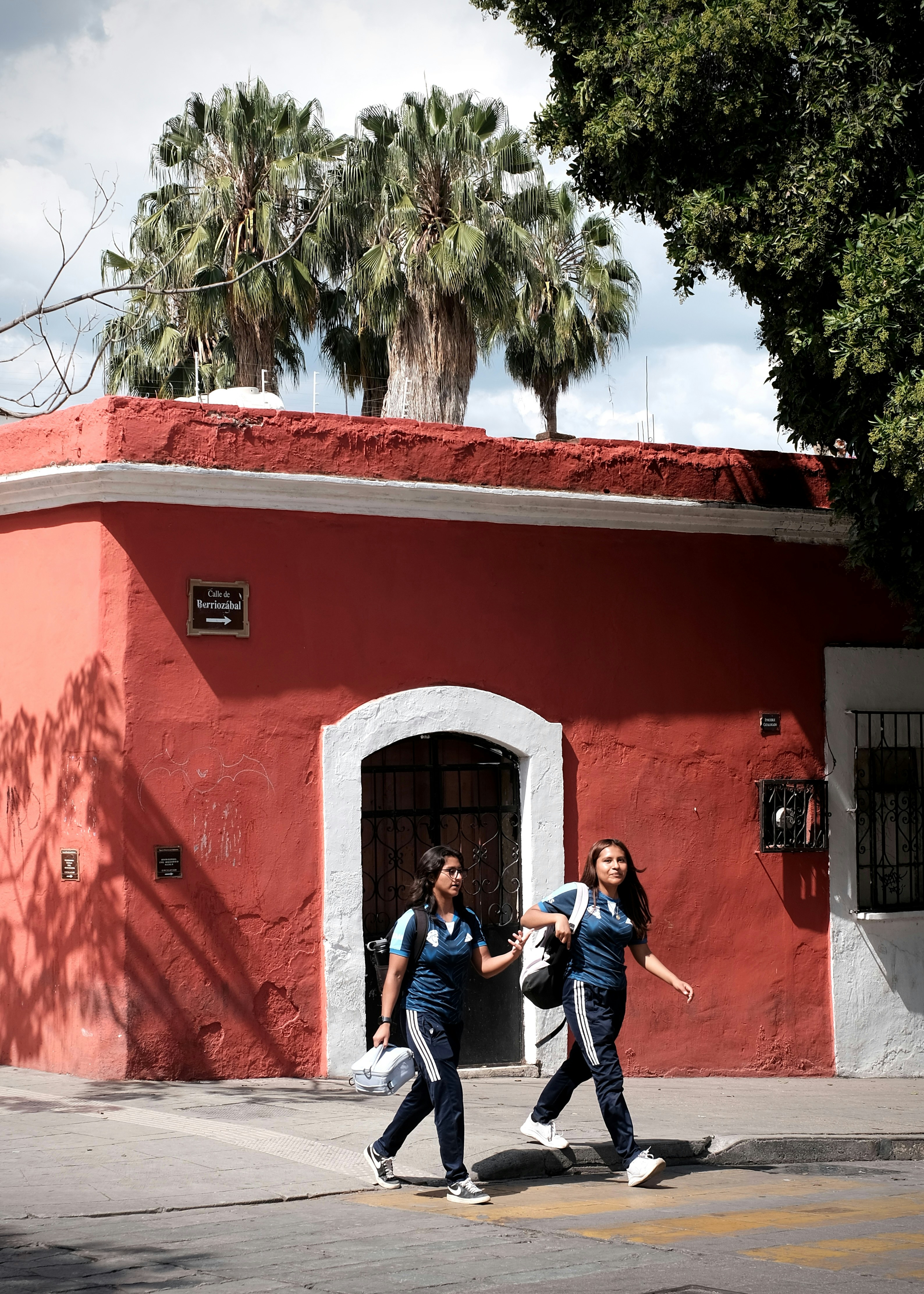 Two schoolgirls stride past a red adobe wall under palm trees