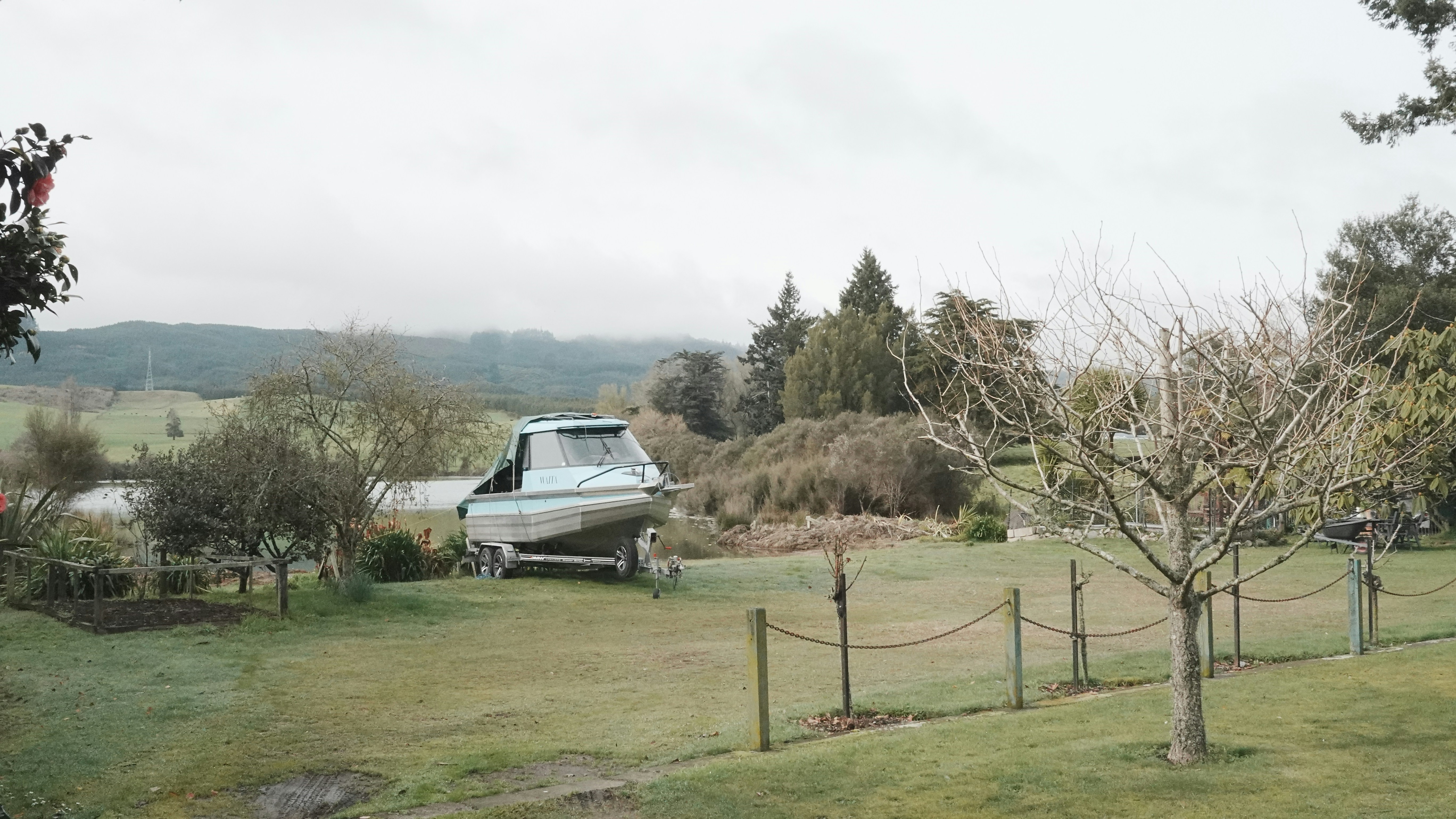 A small boat on a grassy lawn beside a fence and leafless trees, set against a distant rural landscape with rolling hills and a cloudy sky. Note: the scene appears as a garden or park area with the boat as a focal point.