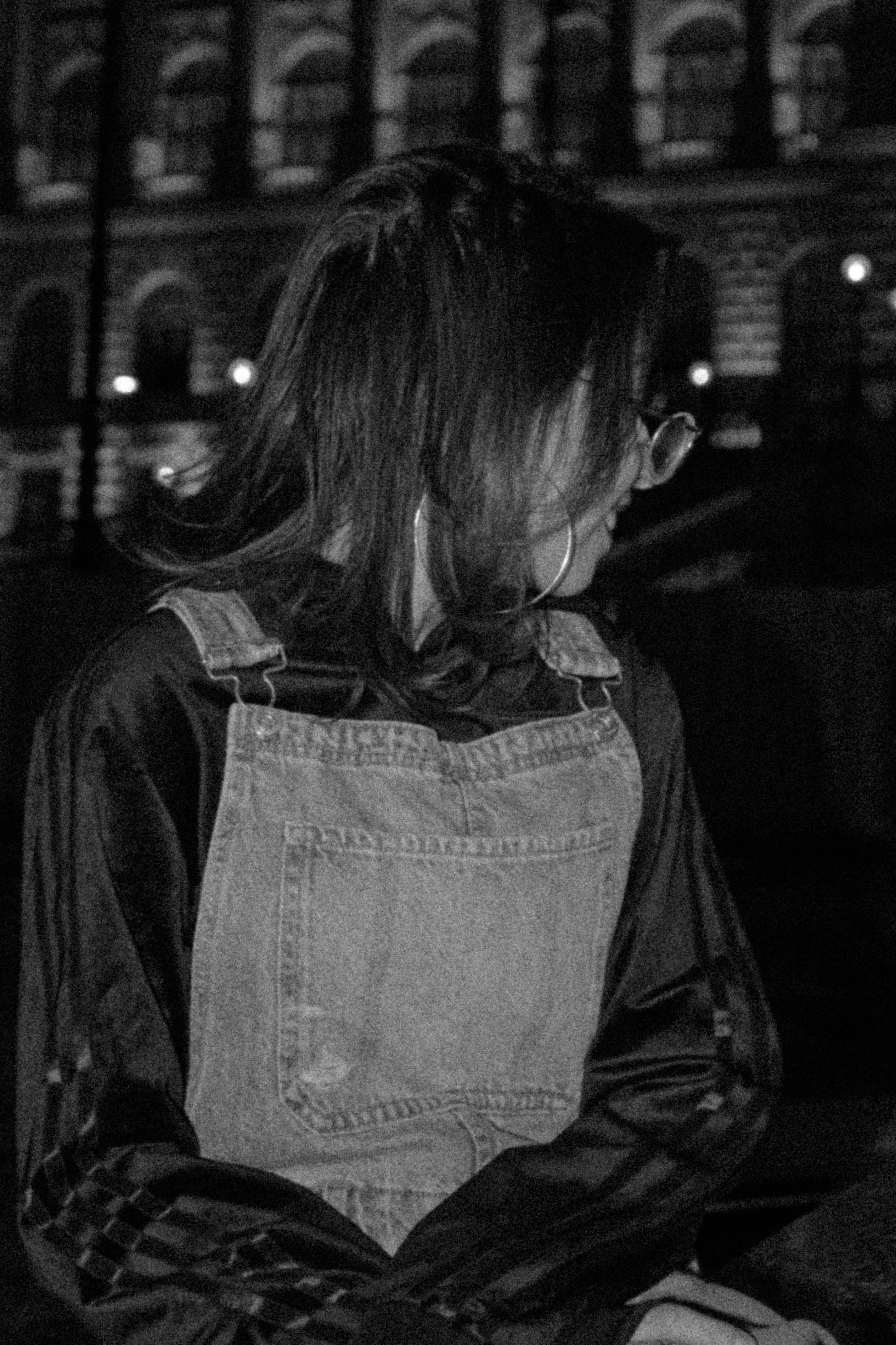 Woman in denim overalls turns away beneath night-lit arches