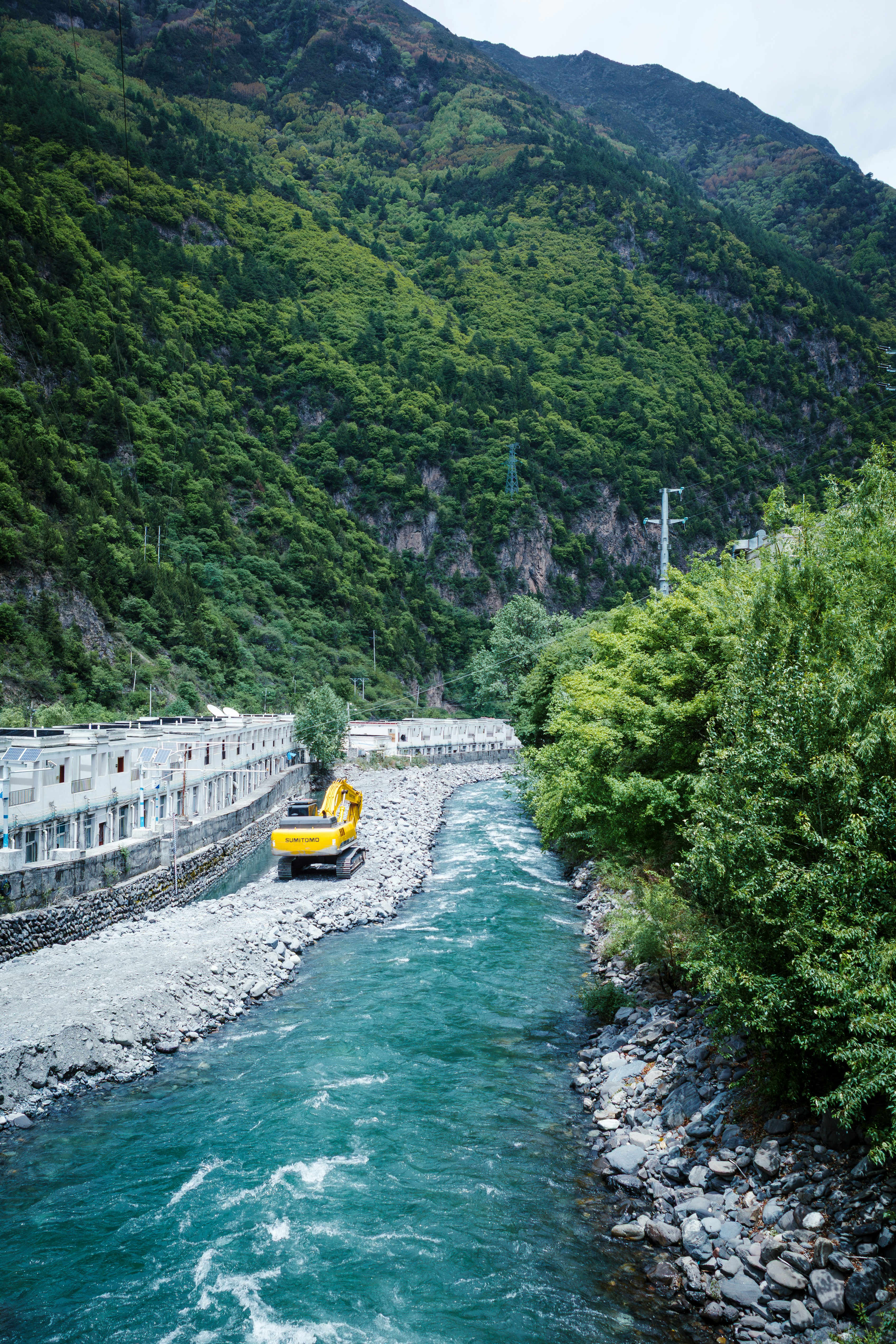 Yellow excavator on a rocky riverbank with mountains.