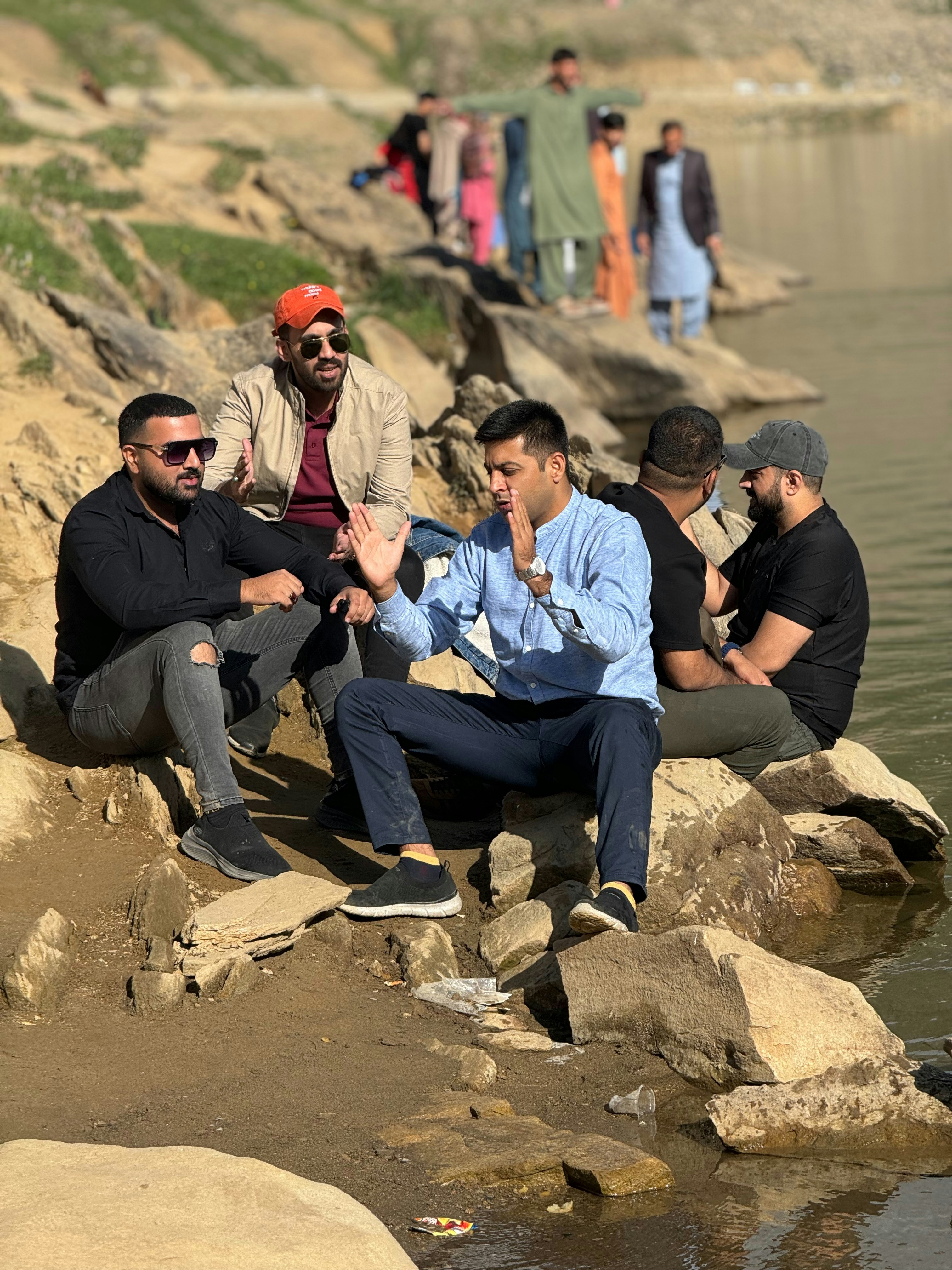 A group of men sit and talk on rocky riverbank ledges by the water’s edge, with several in the background and a distant pathway. The scene captures casual outdoor camaraderie near a lakeside setting.
