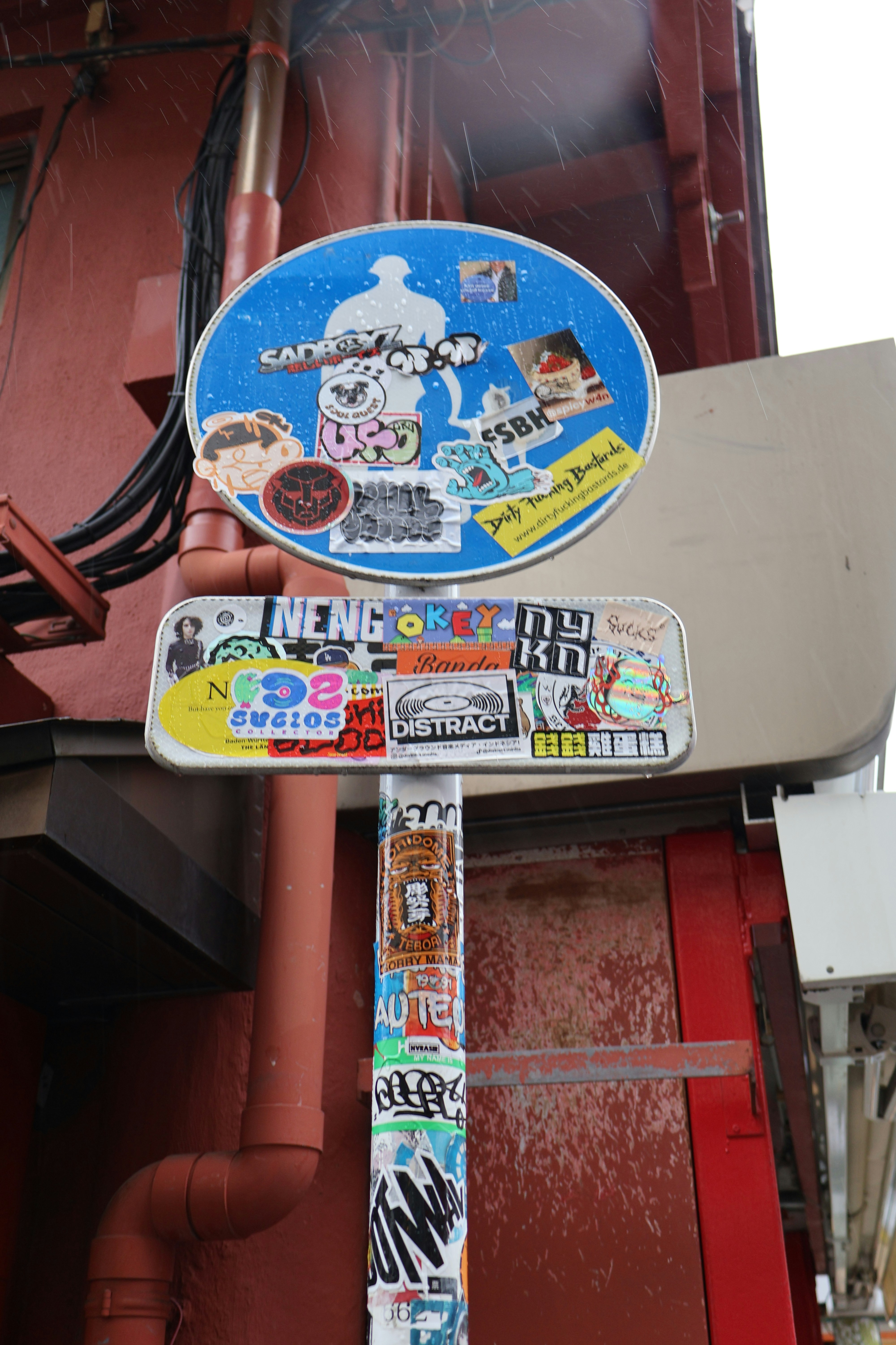 Stickered Street Sign Under Rain on Red Facade