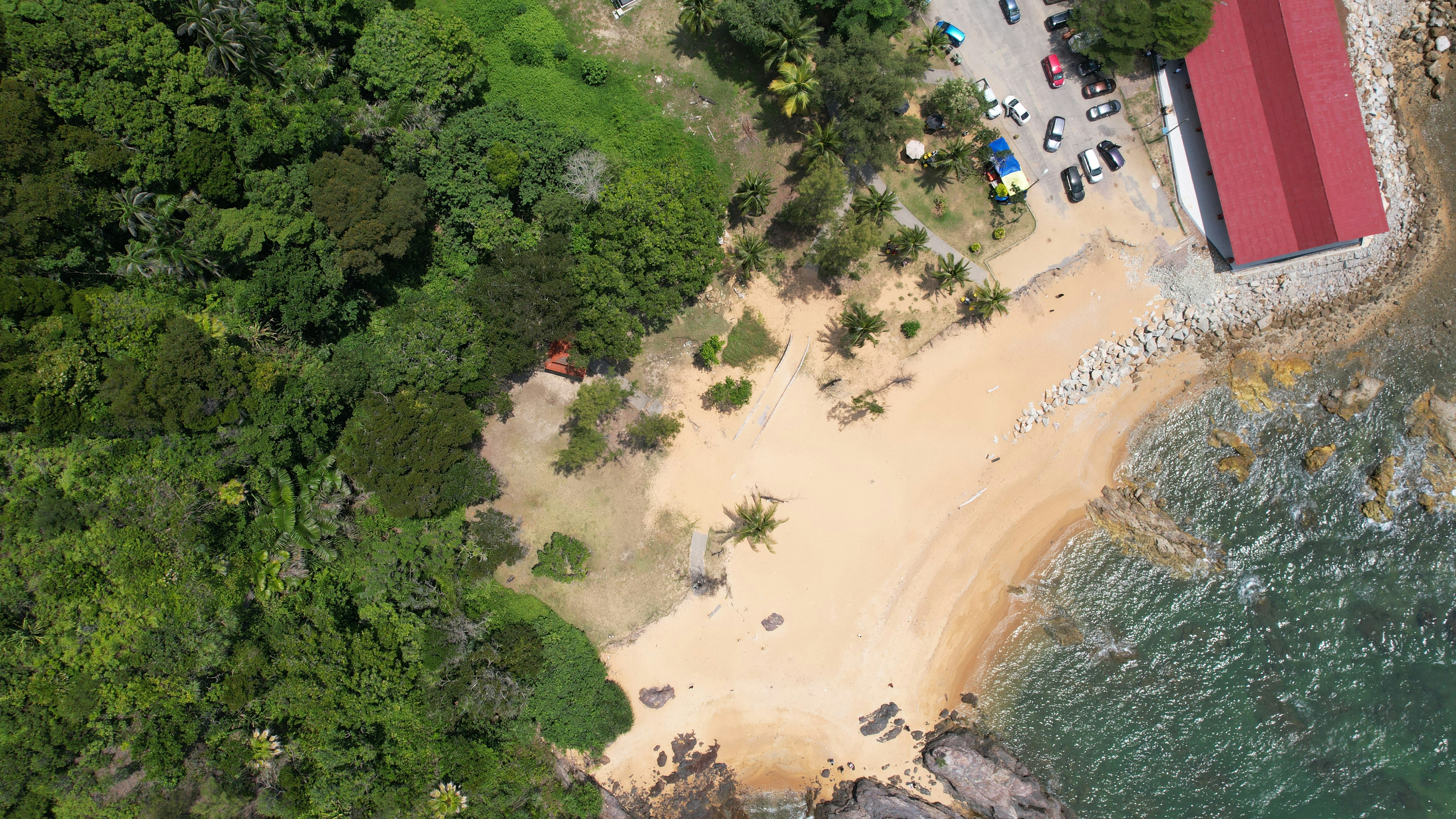 A sandy cove bordered by dense greenery on the left and rocky shoreline on the right, with a red-roofed structure near the water and scattered vehicles along the shore.