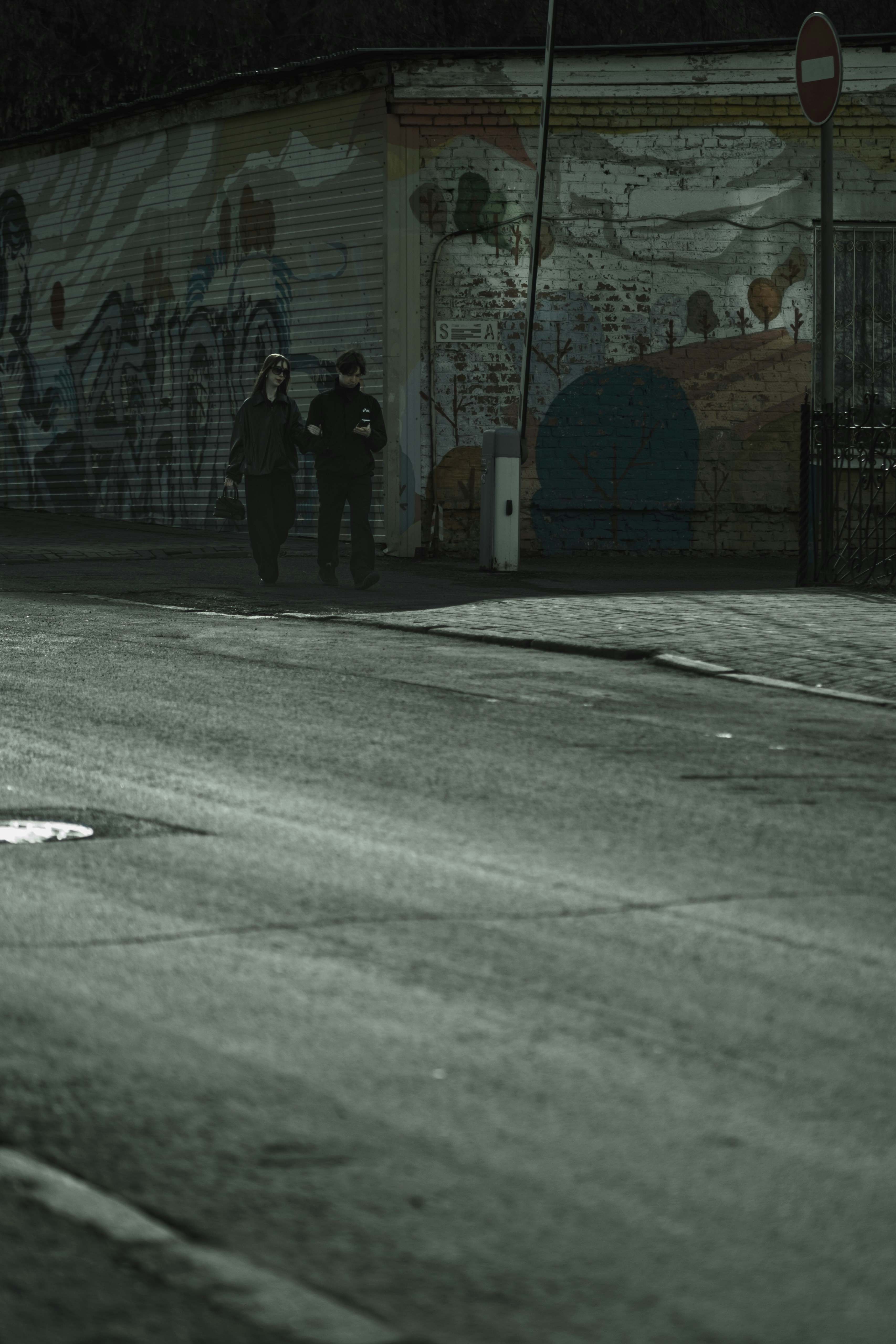 Two pedestrians in shadow along graffiti wall beneath a curved streetlamp