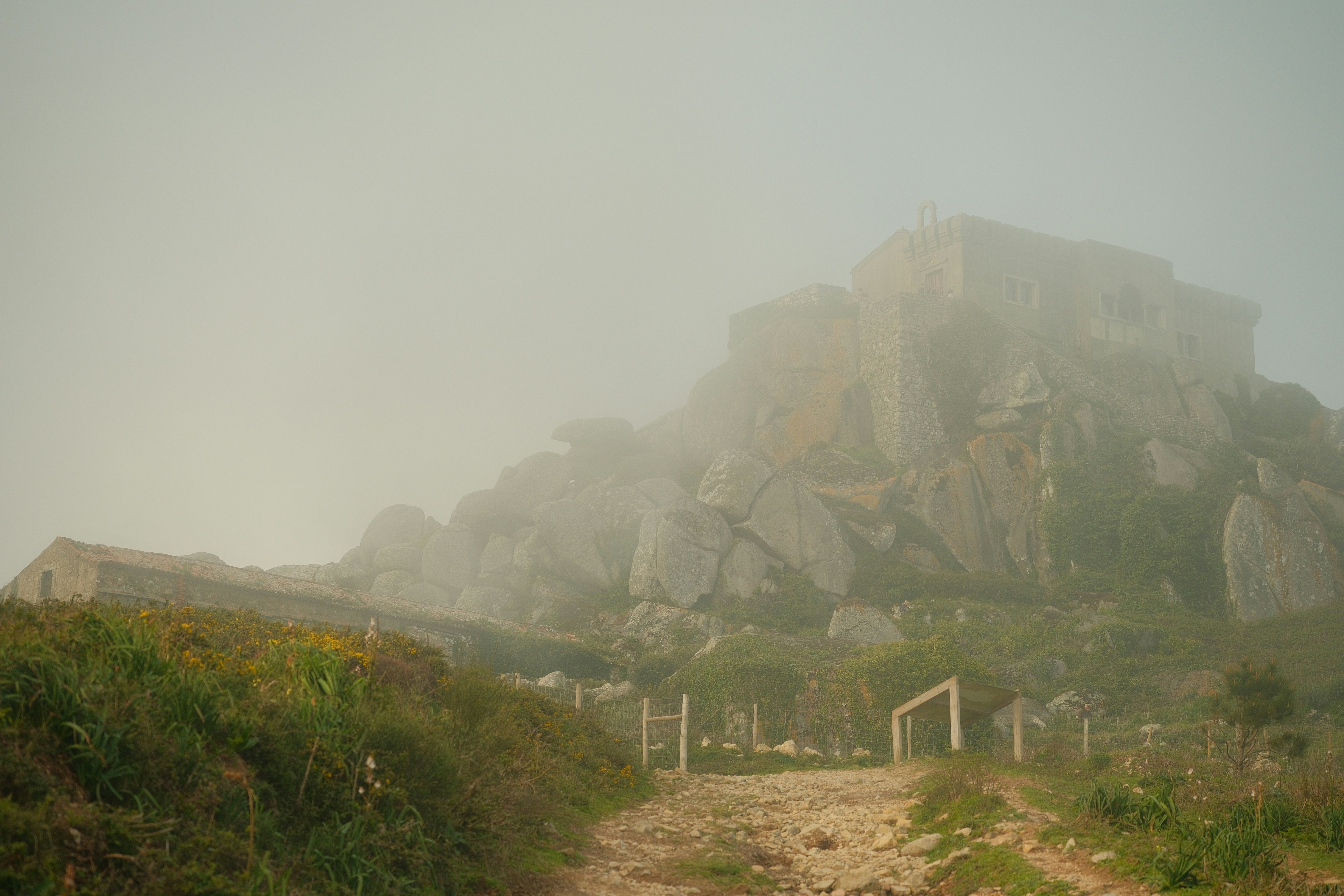 The Peninha Monastery emerges through dense fog atop the Sintra mountains, surrounded by rugged boulders and lush greenery, creating a quiet and atmospheric scene. // O Mosteiro da Peninha surge envolto em nevoeiro no topo da serra de Sintra, rodeado por grandes rochas e vegetação densa, criando uma atmosfera serena e misteriosa.