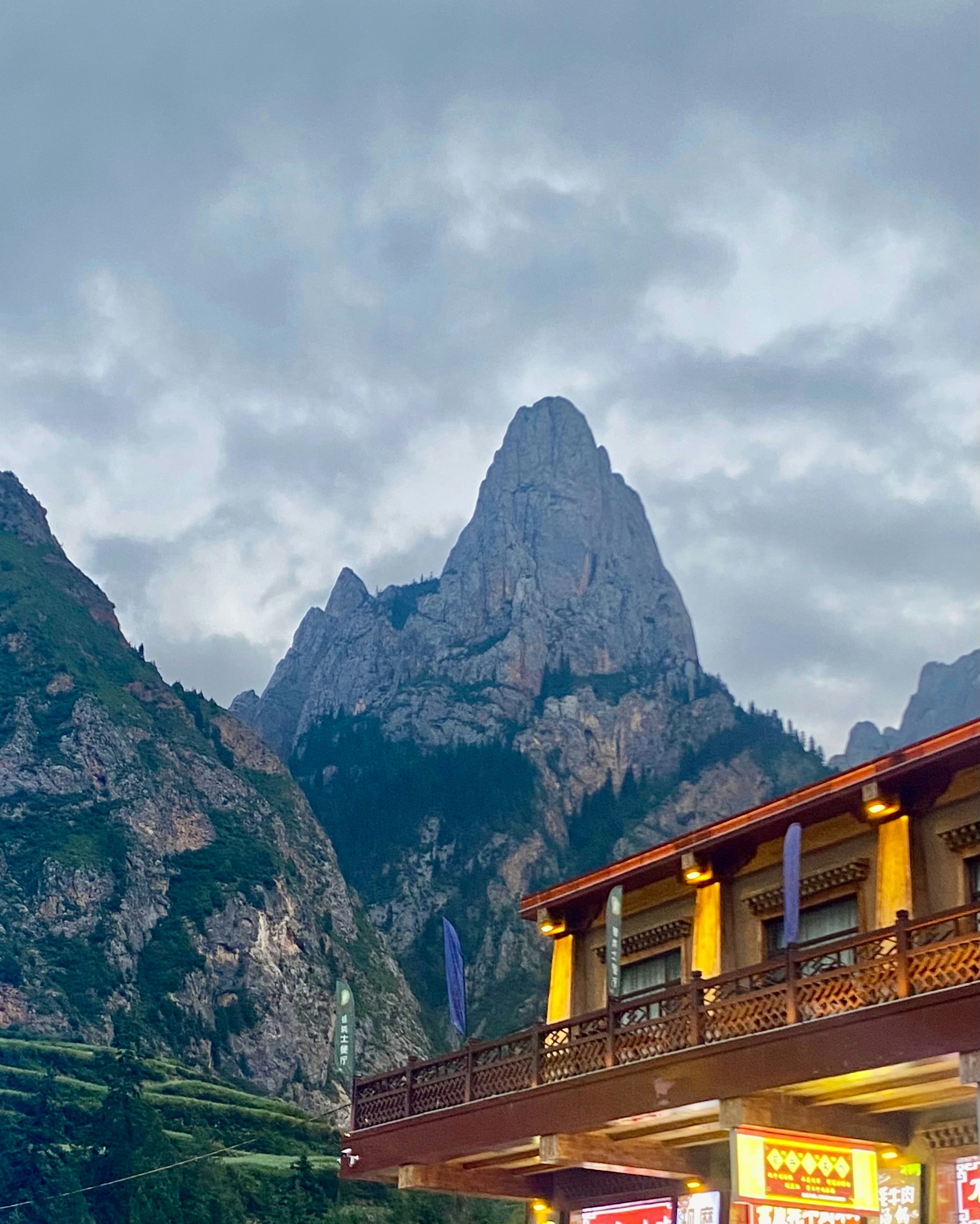 Snow-dusted peak rises behind a wood-and-stone balcony of a mountain lodge, with cloudy sky overhead.