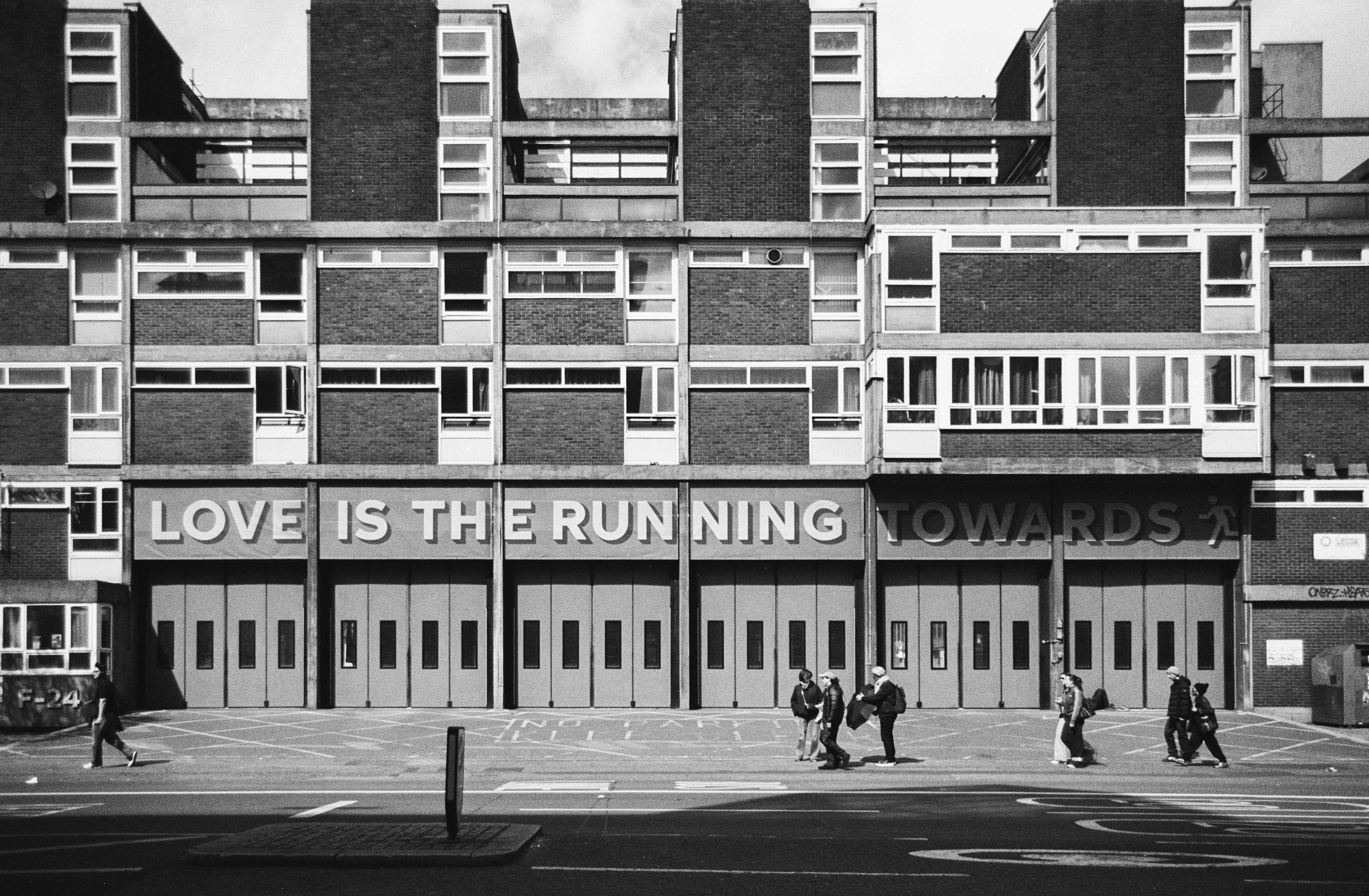Black and white street photograph of a large typographic mural reading “Love is the running towards”, set against a modernist building facade. Shoreditch High Street, London
