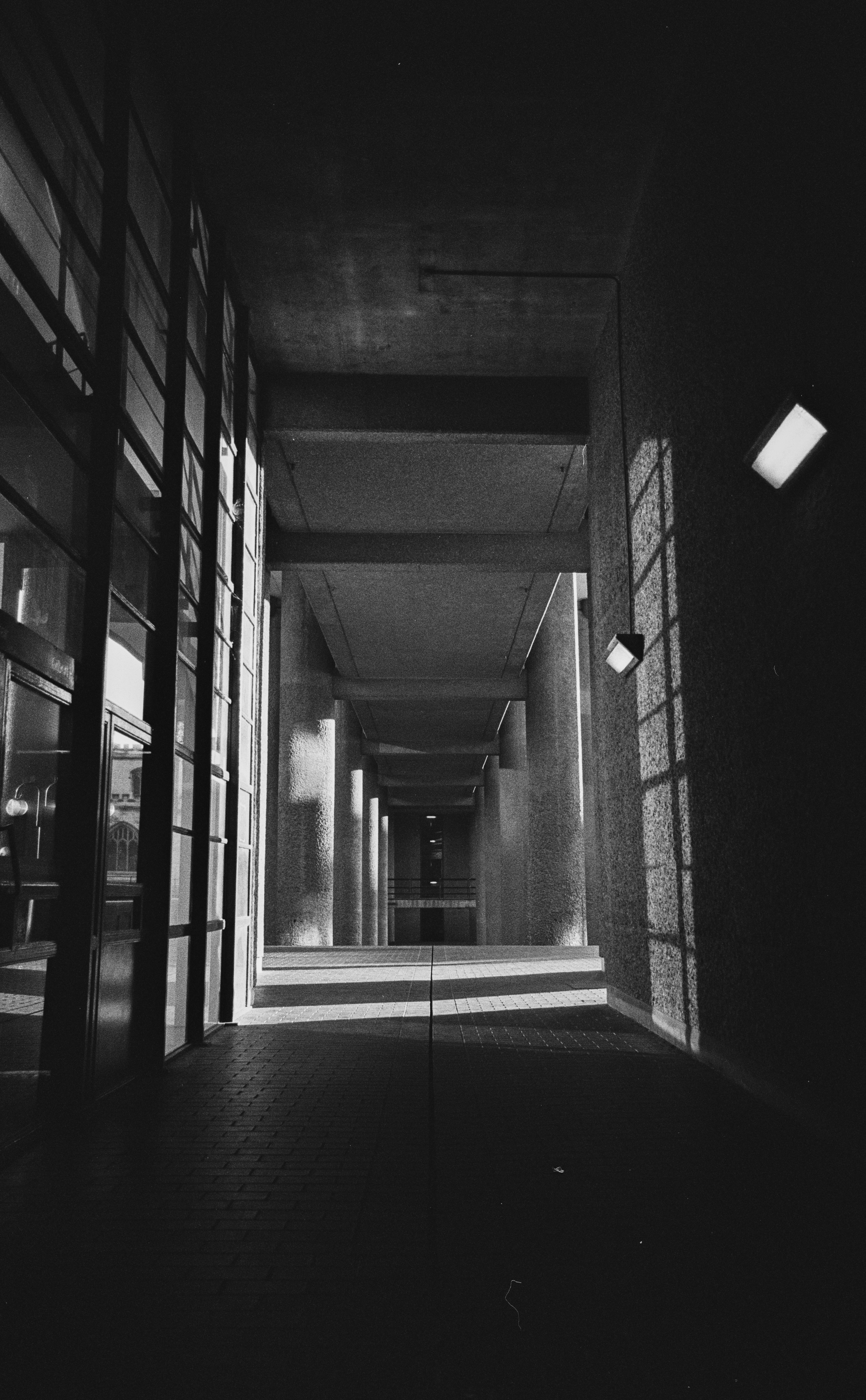 Black and white architectural photograph of a dark covered walkway, in Barbican, London, with strong contrast between shadow and daylight
