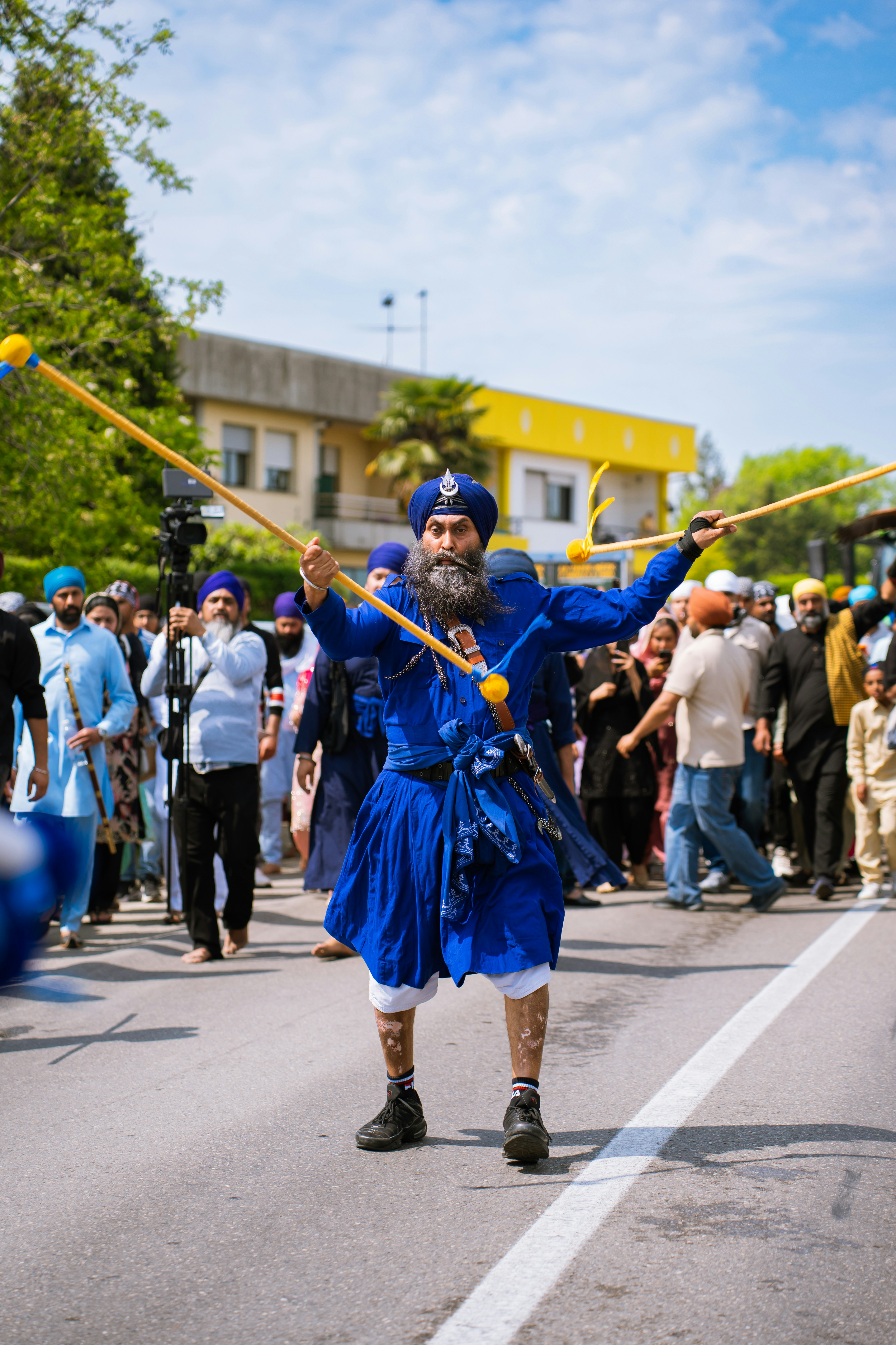 Blue-clad martial artist leads a street procession with extended spear staves