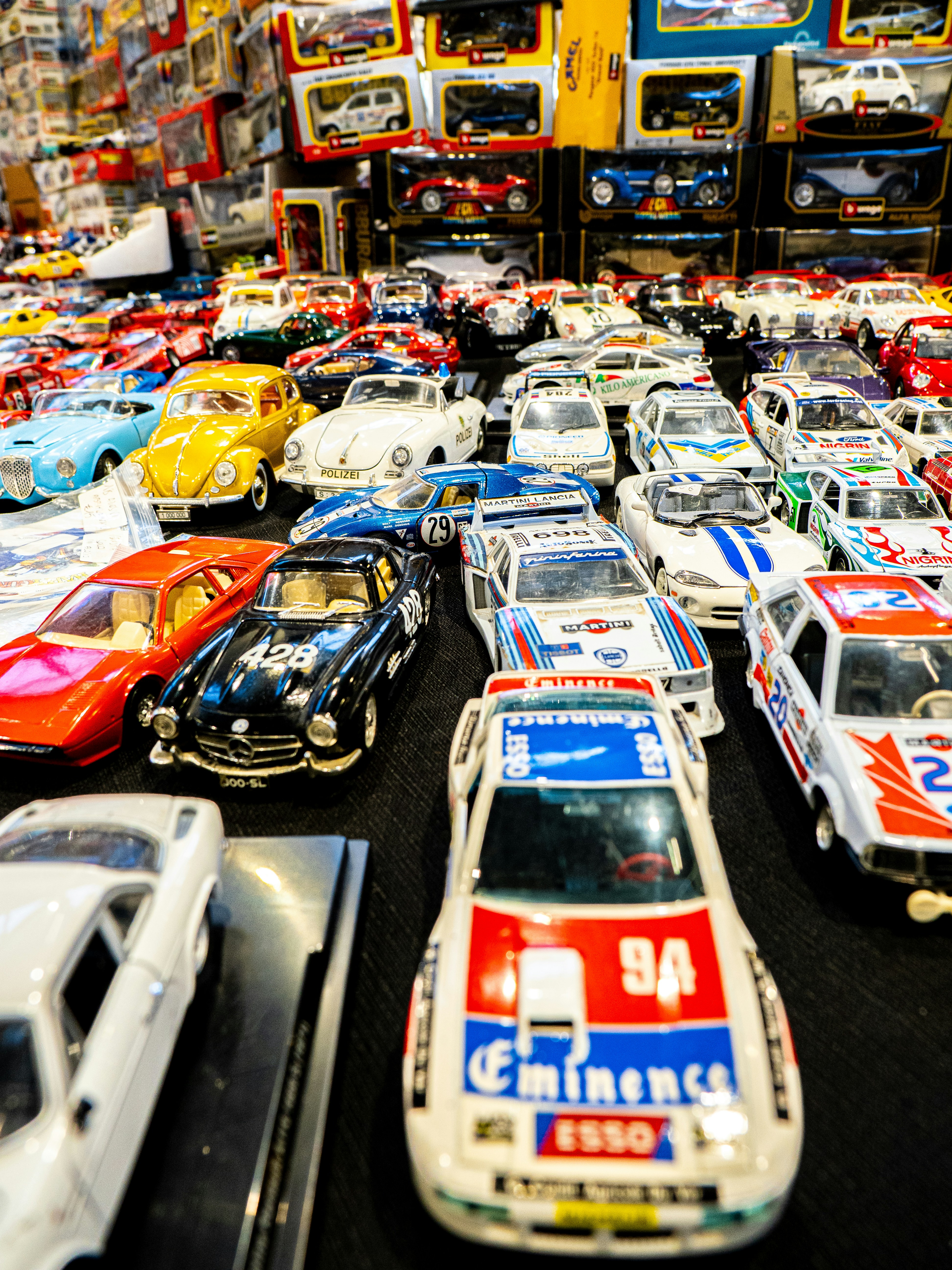 A dense display of colorful toy race cars arranged on a black surface with packaging and boxes in the background.