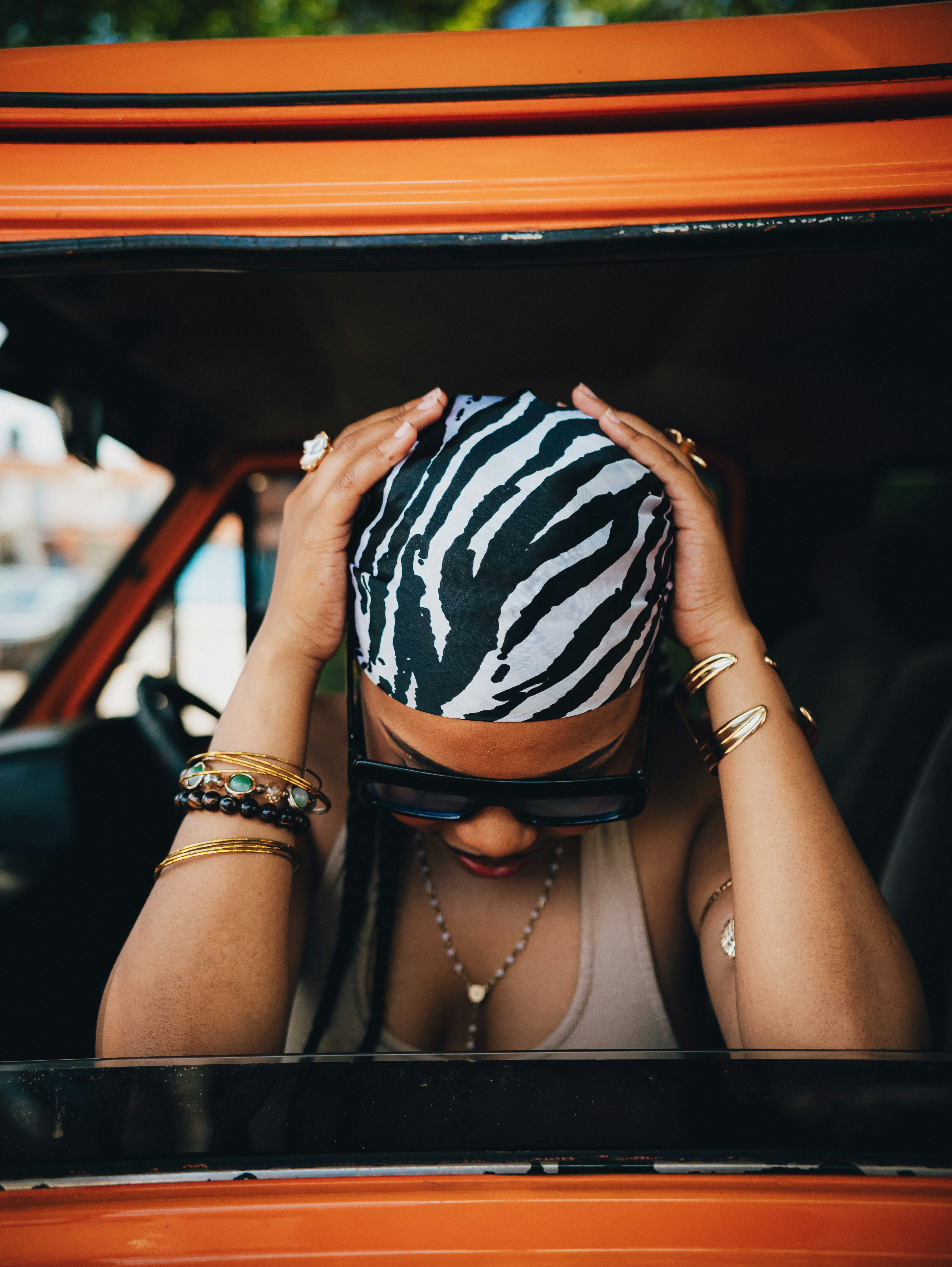 A person leans forward in an orange vehicle, adjusting a zebra-pattern bandana over their head while wearing dark sunglasses and layered jewelry; the scene is framed by the car window and interior.