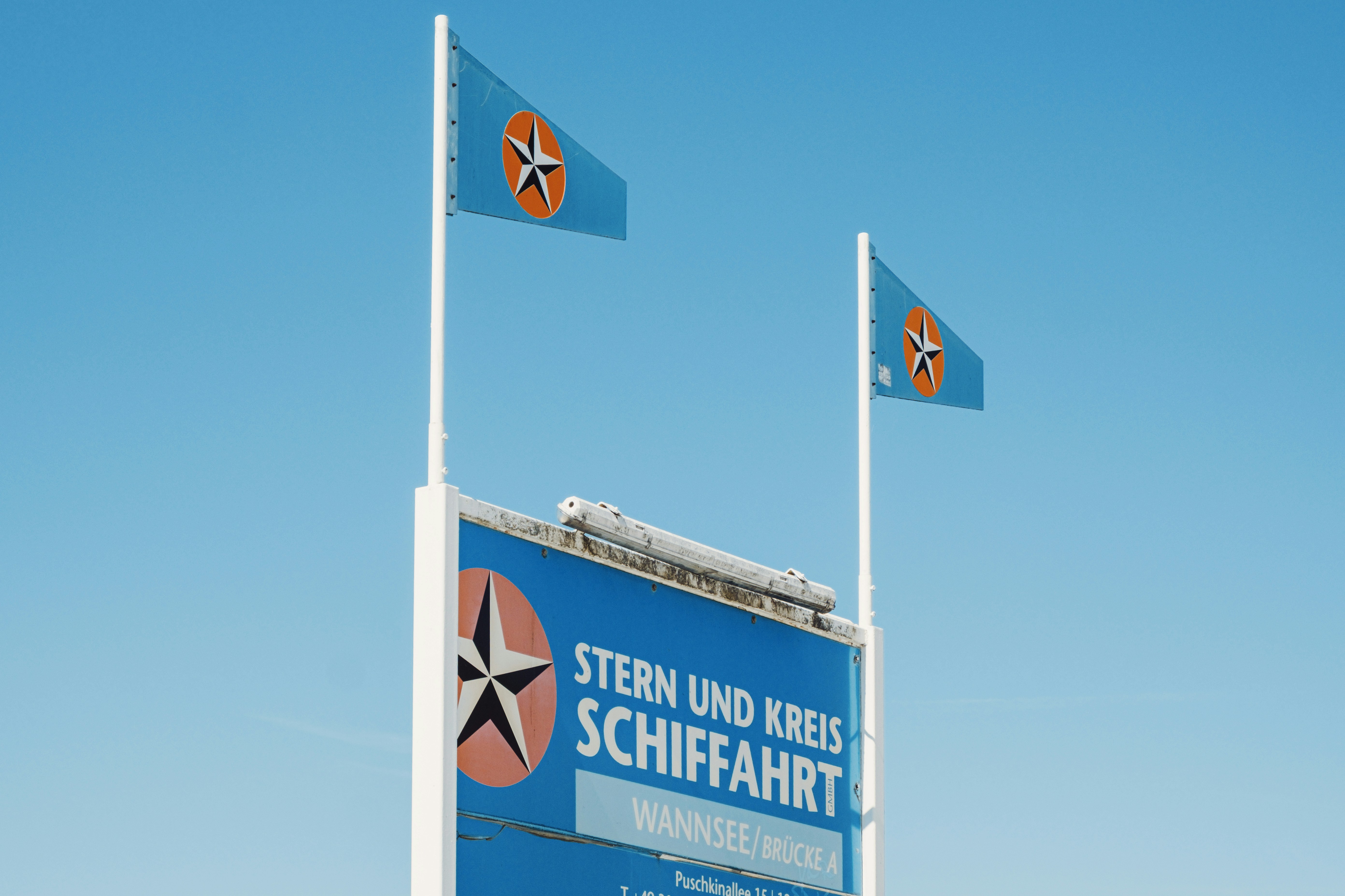 Blue sign with white text and star emblem, flanked by two flagpoles against a bright blue sky.