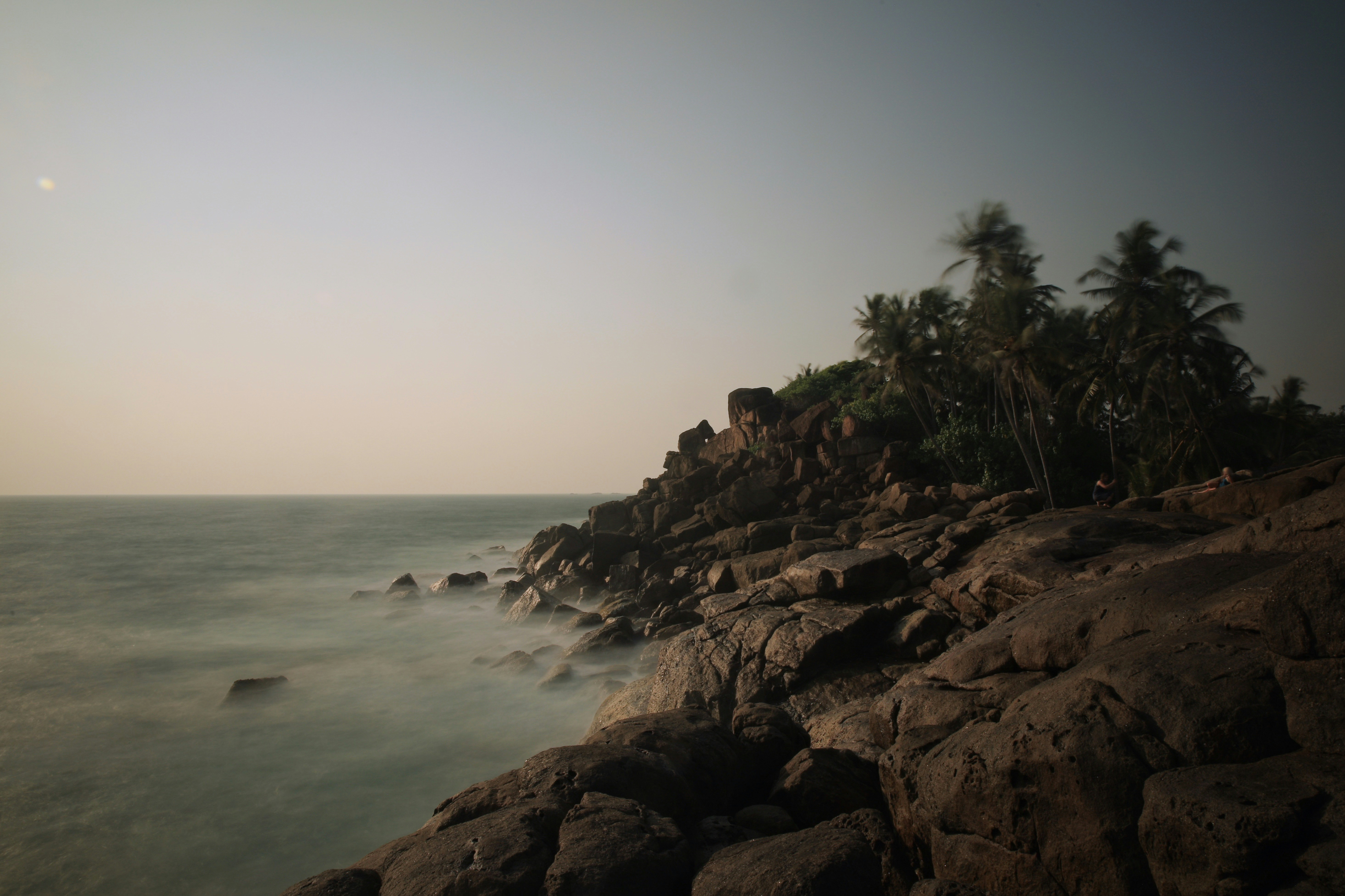 Rocky shoreline at sunset with palm silhouettes and distant horizon