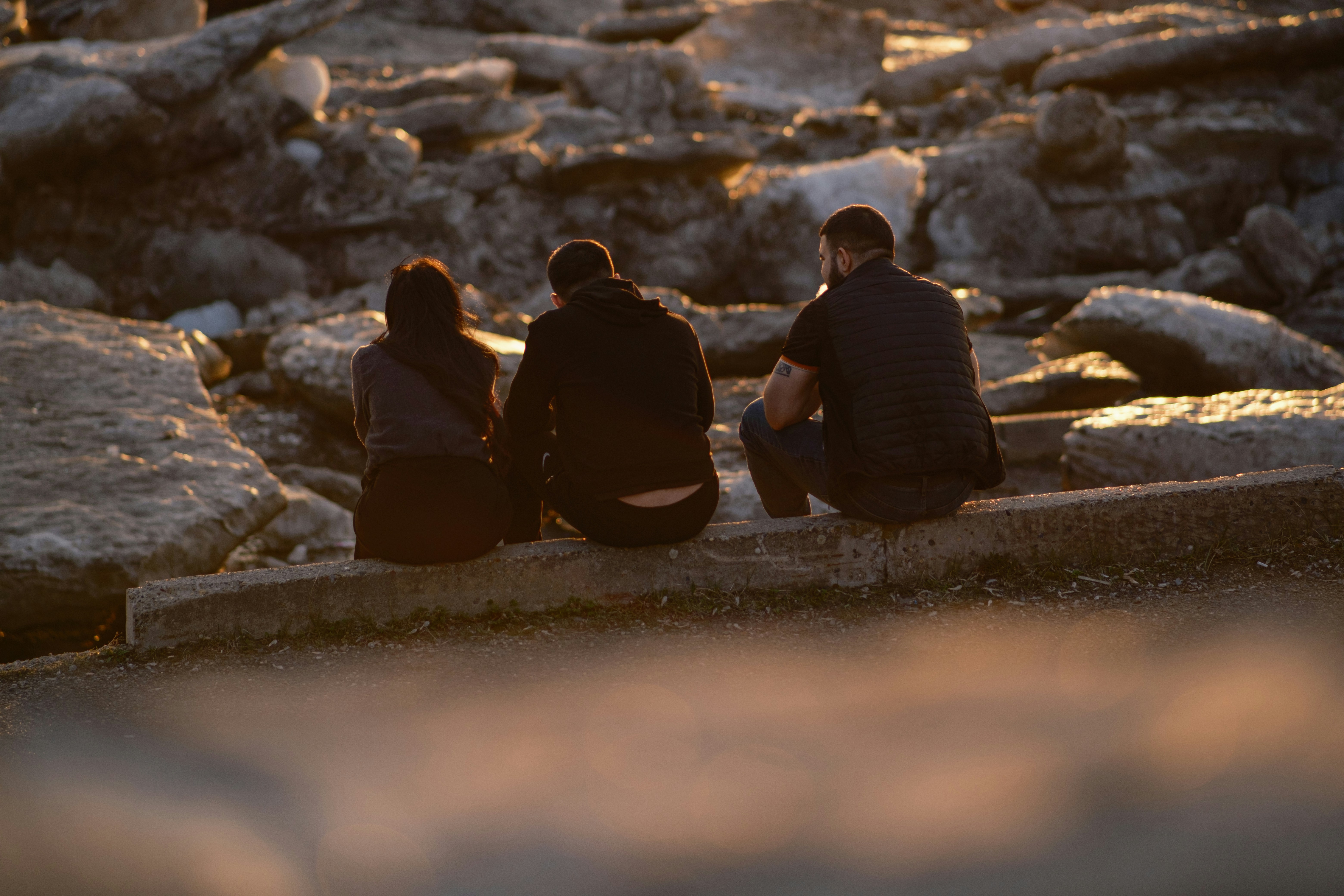 Three silhouettes on a sunlit seawall watching the rocky shoreline