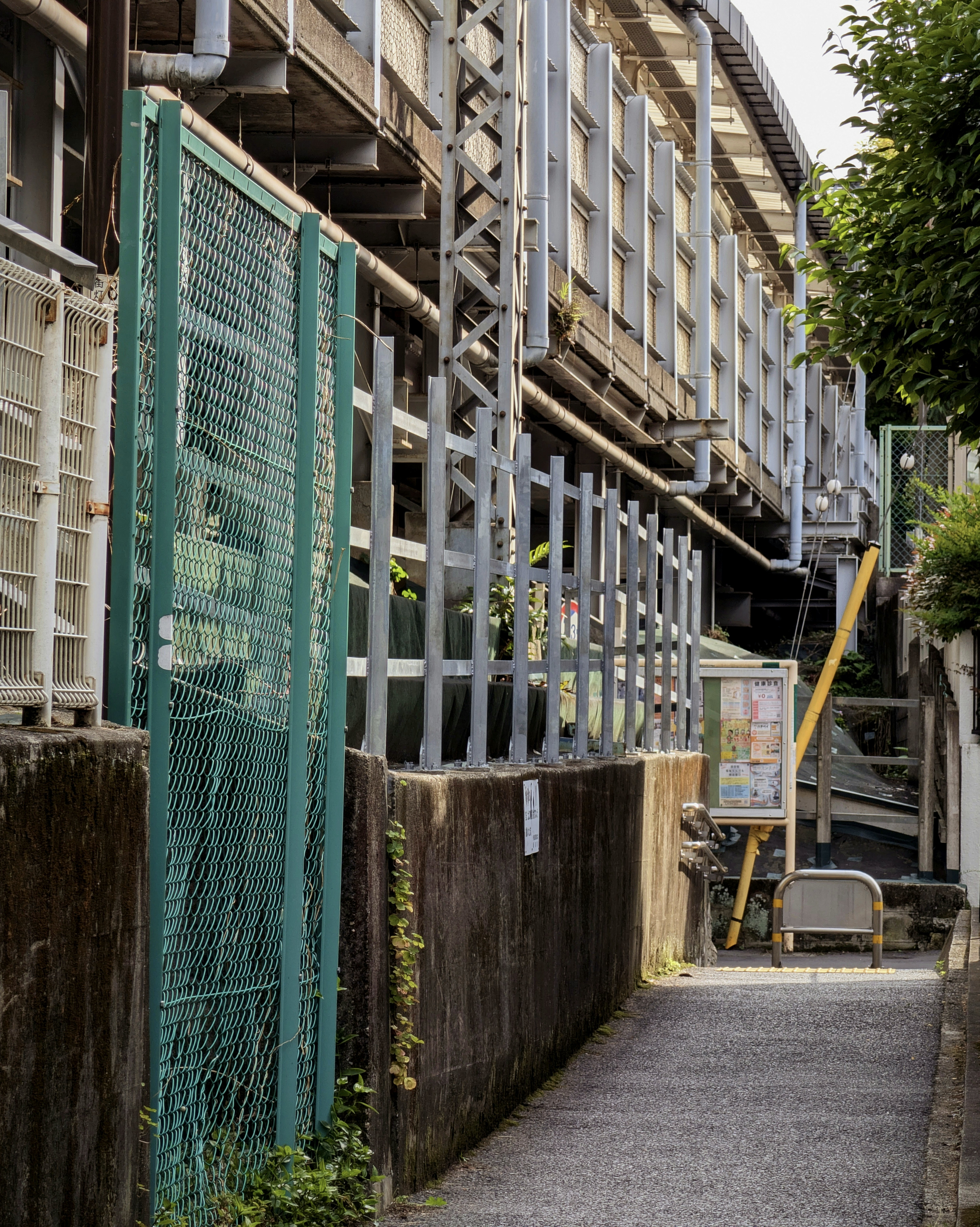 Narrow alley with concrete walls and green chain-link fence, a utility structure overhead, and a small posted board at the far end.