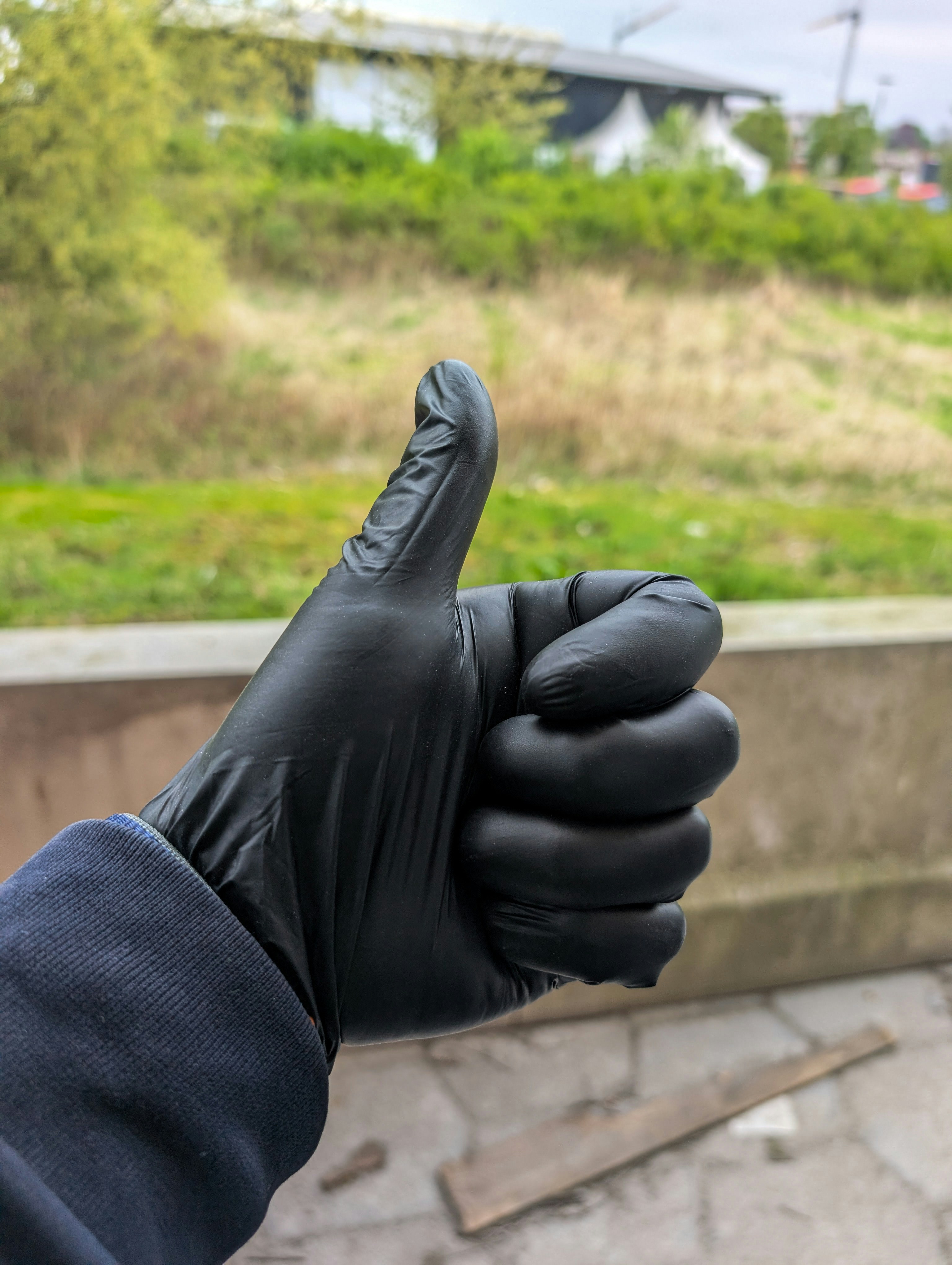 Close-up of a gloved hand giving a thumbs-up in the foreground with an out-of-focus urban-parkside background and concrete foreground.