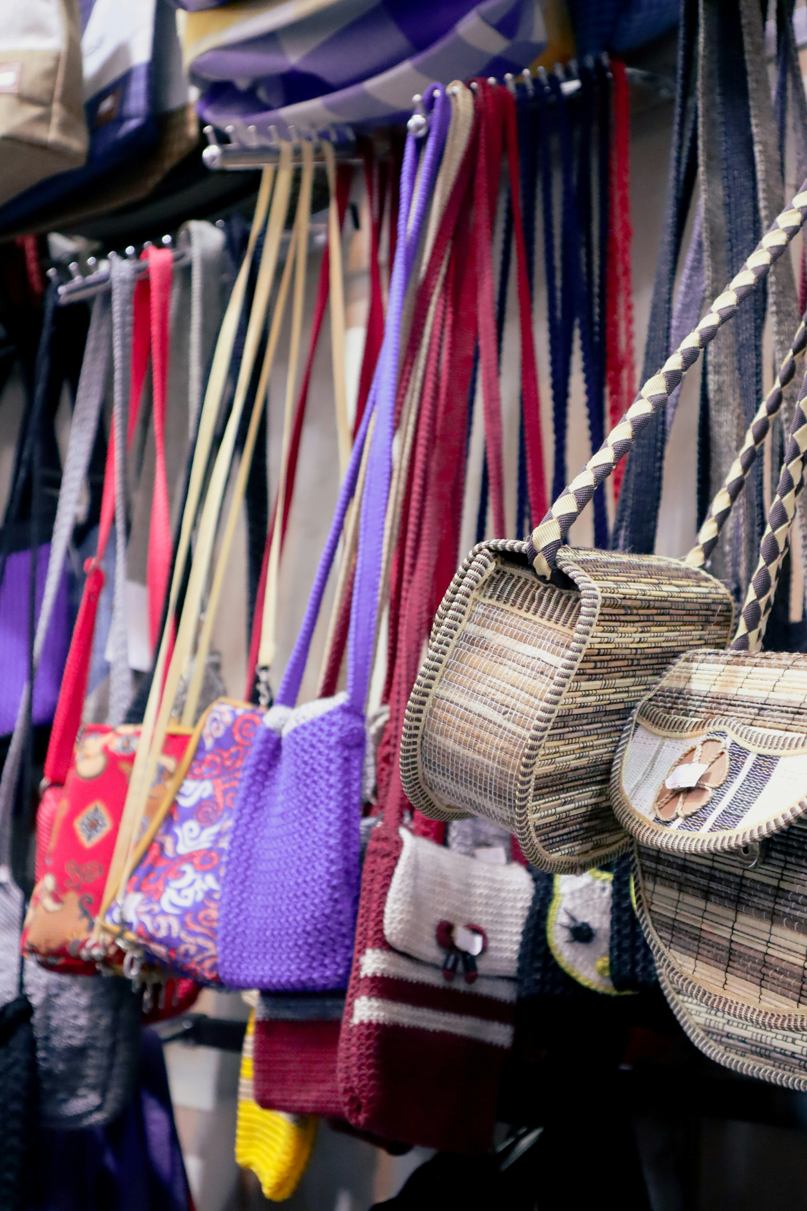 Row of woven sling bags and purses hanging in a market stall, with varied textures and colors against a blurred background.