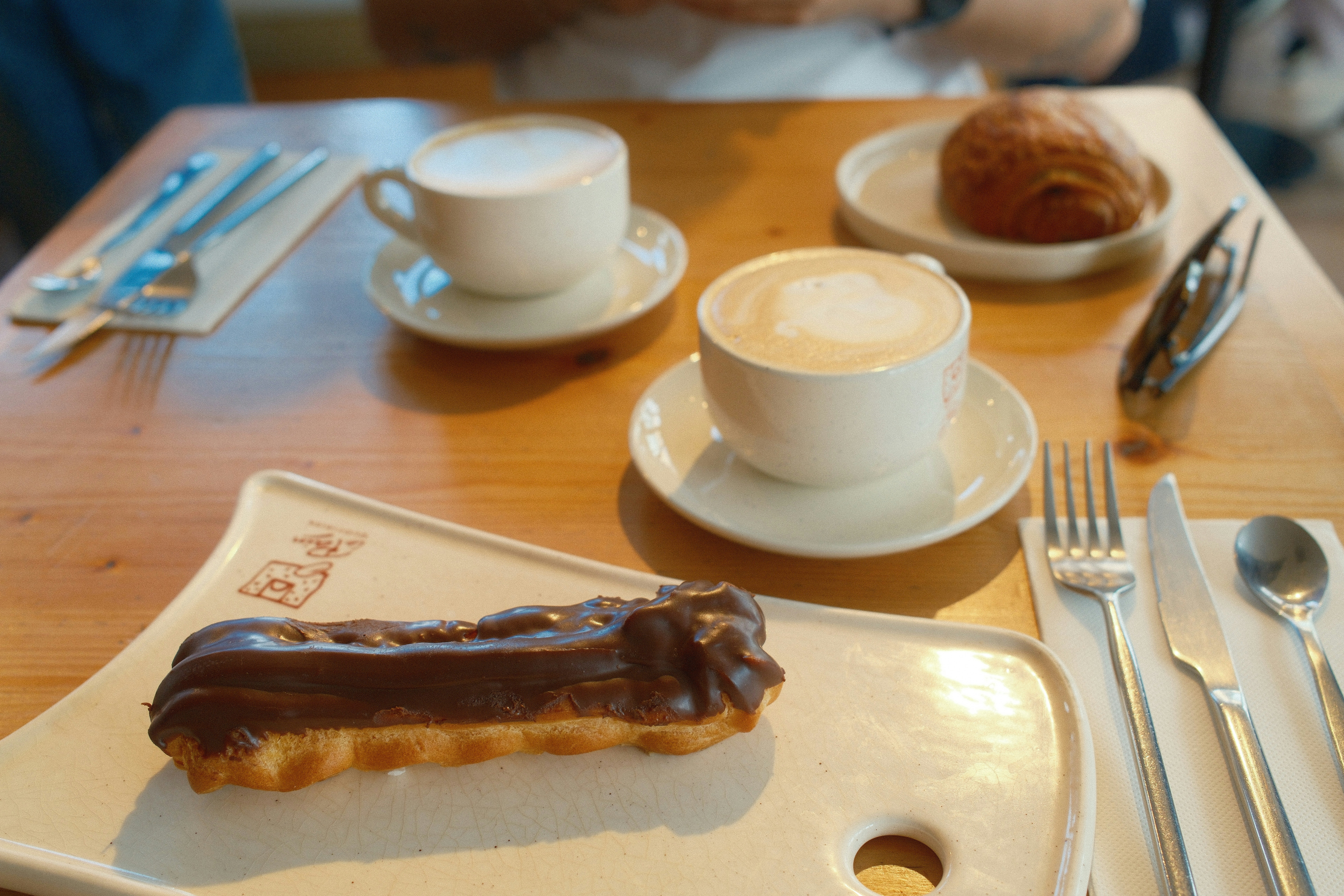 Café scene with a chocolate éclair in the foreground, flanked by two cups of coffee and a croissant in the background on a warm wooden table.