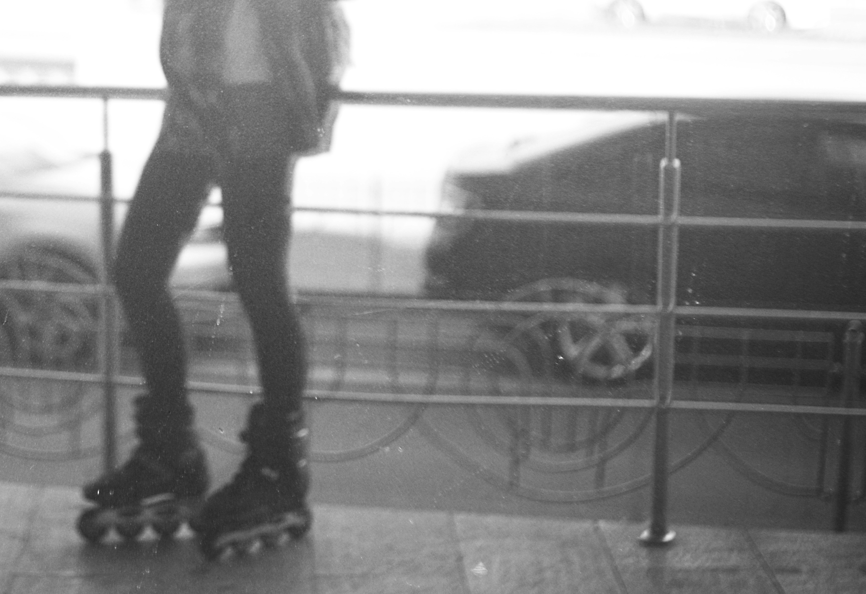 Grayscale scene of a person on inline skates leaning against a railing, background cars blurred in motion; a street-side moment focused on silhouette and movement.