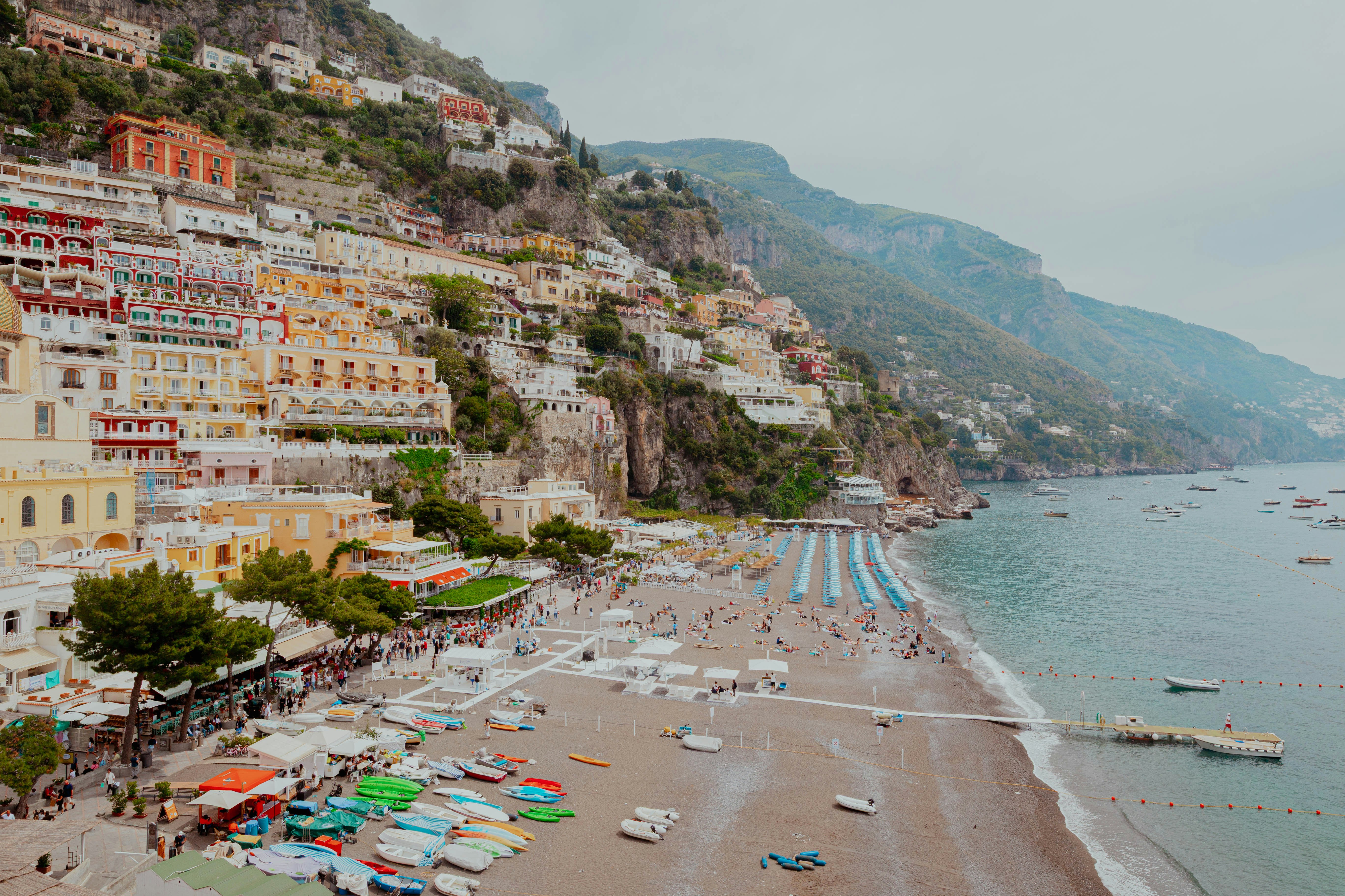 Sunlit beach scene with colorful hillside houses and boats along a curved coastline.