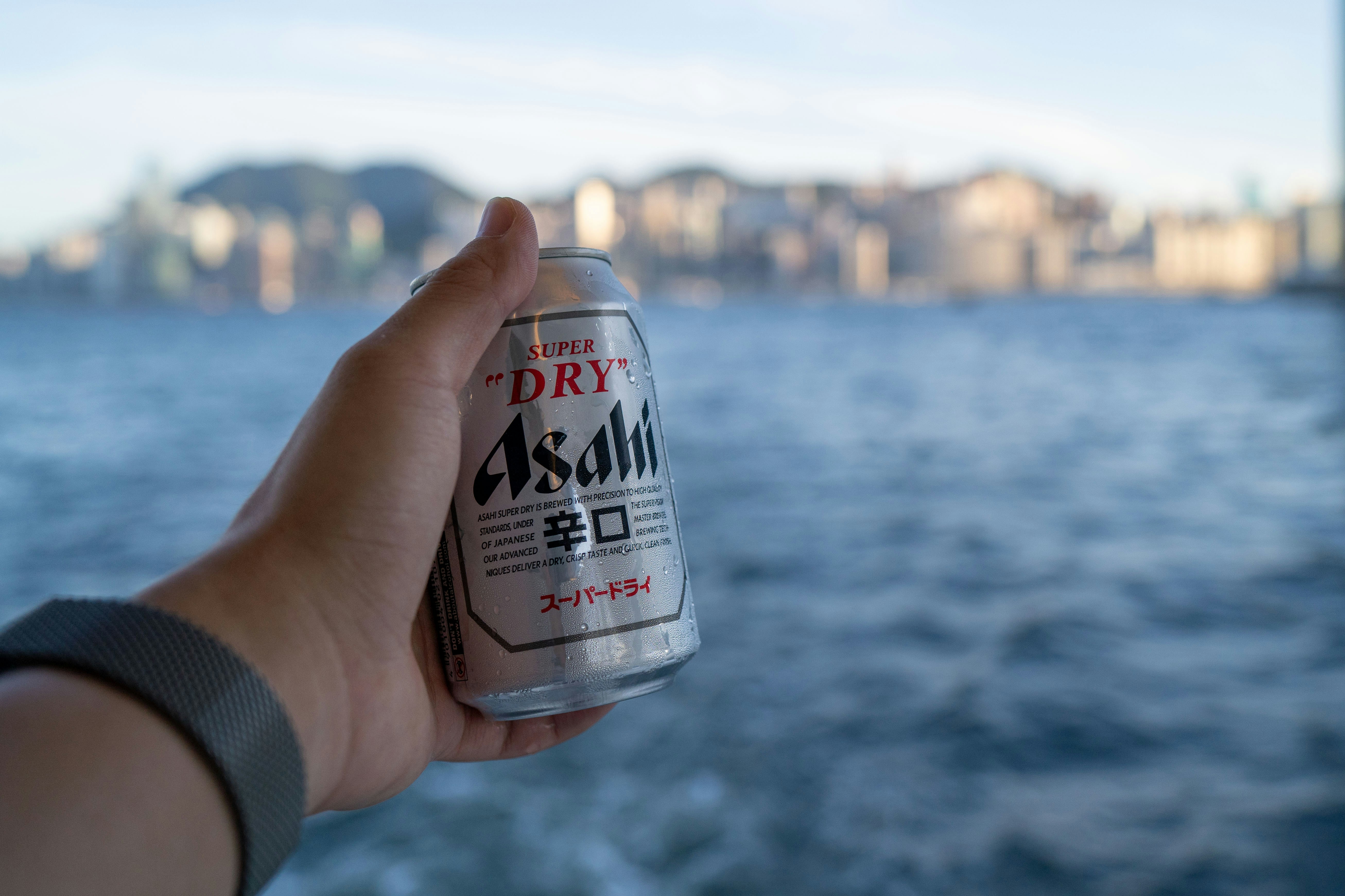Close-up of a hand gripping a can of Asahi beer with a blurred cityscape and water in the background.