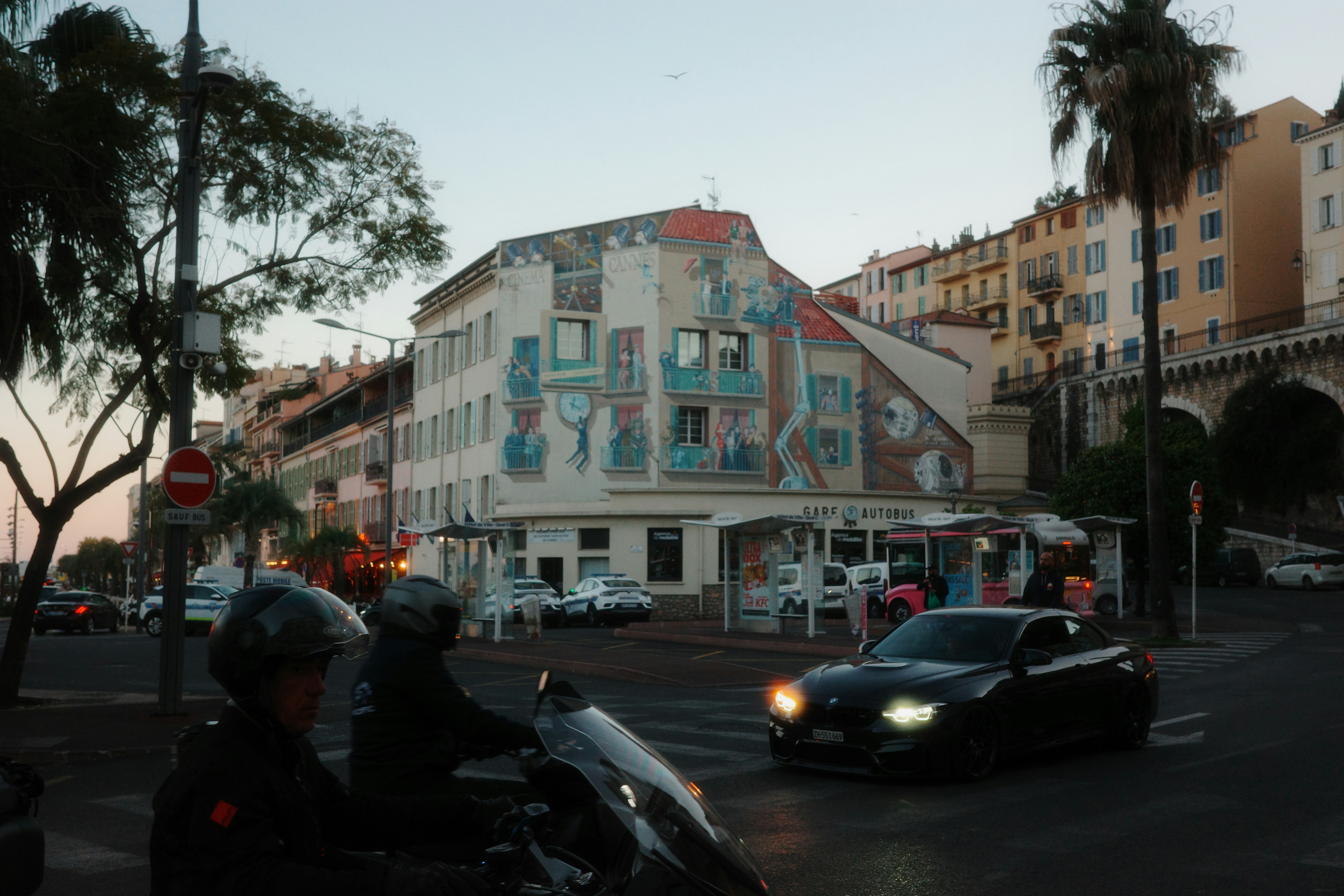 City mural at dusk: street corner with passing cars and scooters